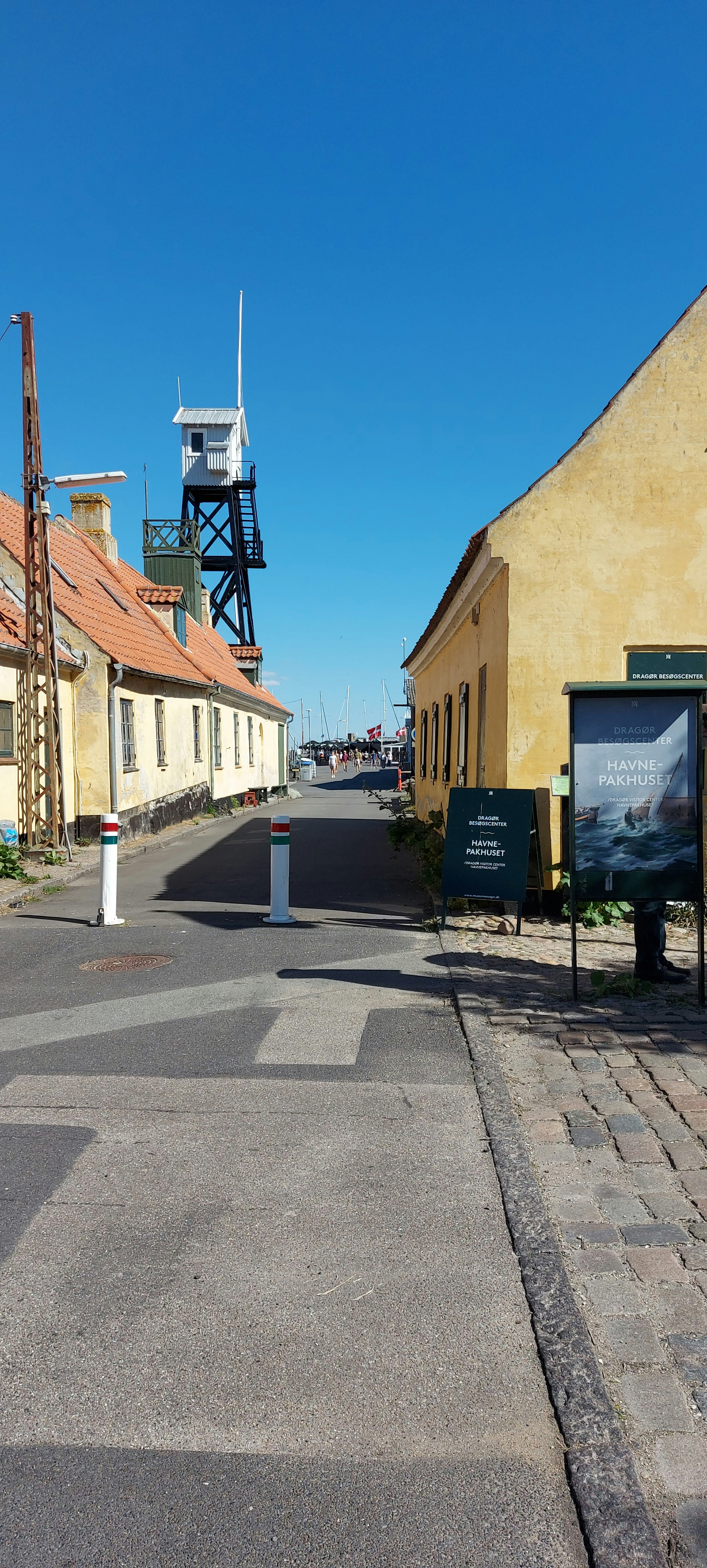 Charming coastal pathway lined with yellow buildings and a lookout tower, leading towards a marina filled with boats.