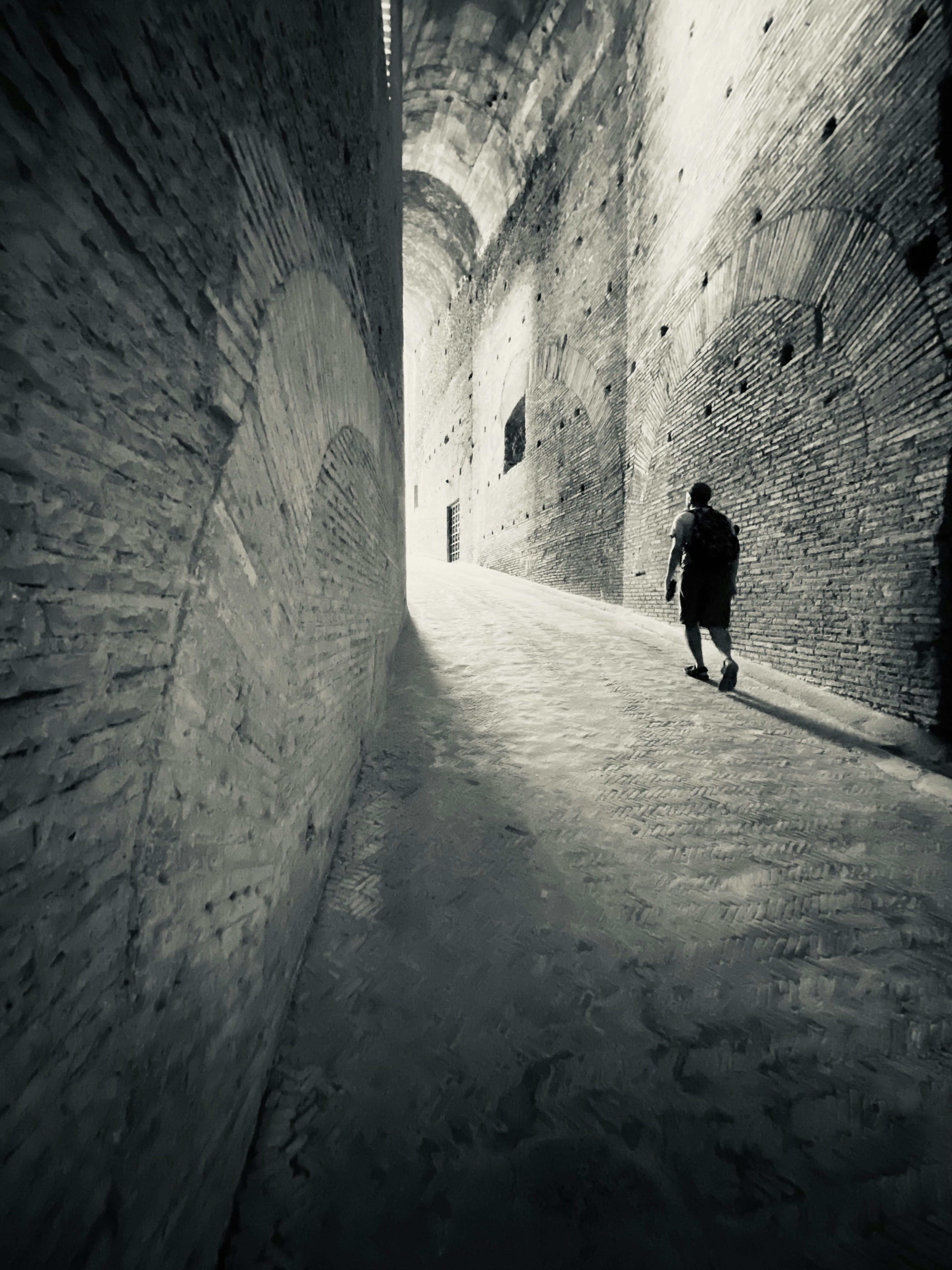 A solitary figure walks through a narrow, dimly lit corridor of ancient brick walls, evoking a sense of history and solitude.
