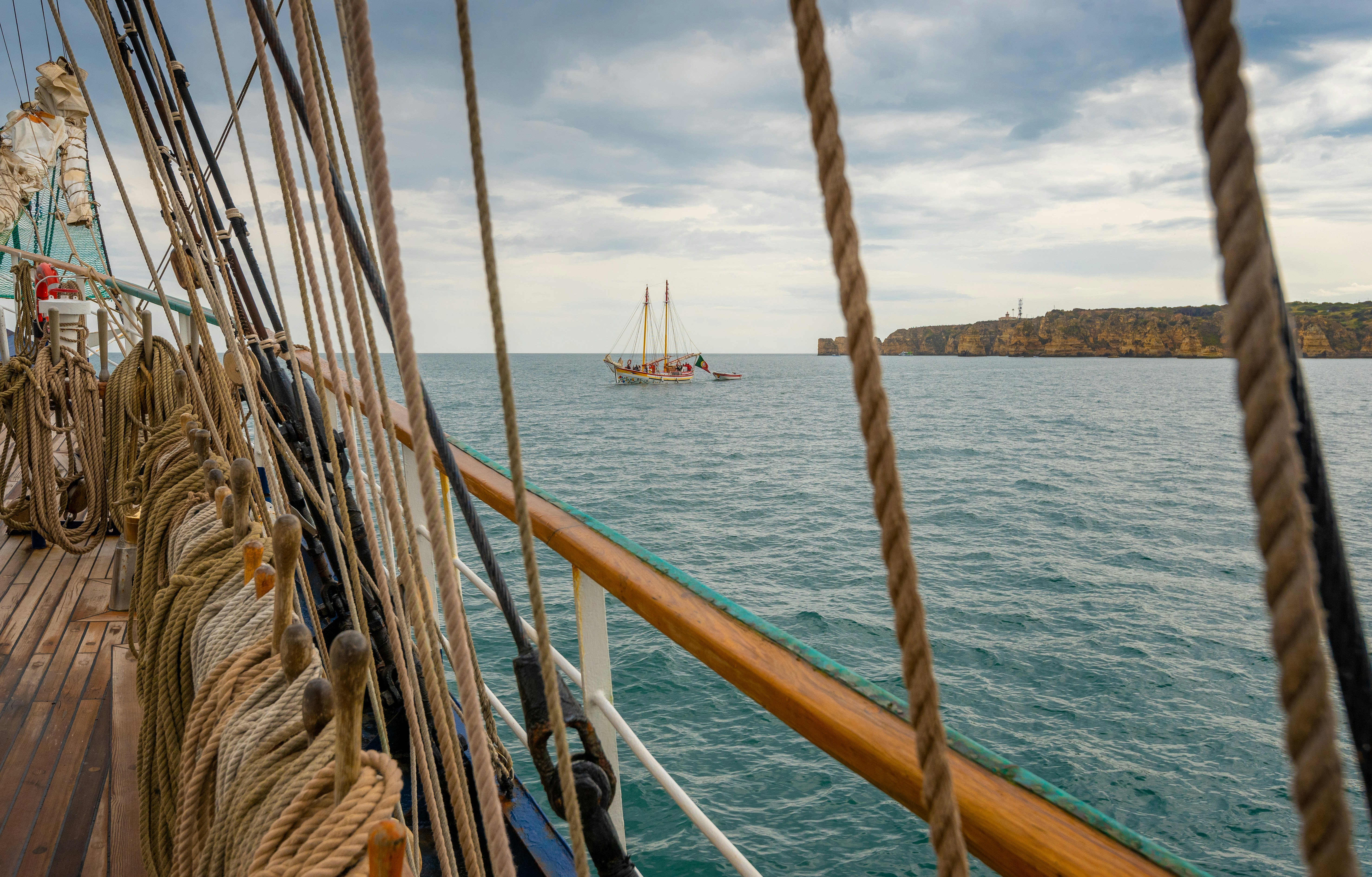 View from a sailing ship deck with ropes in the foreground and distant cliffs under a cloudy sky.