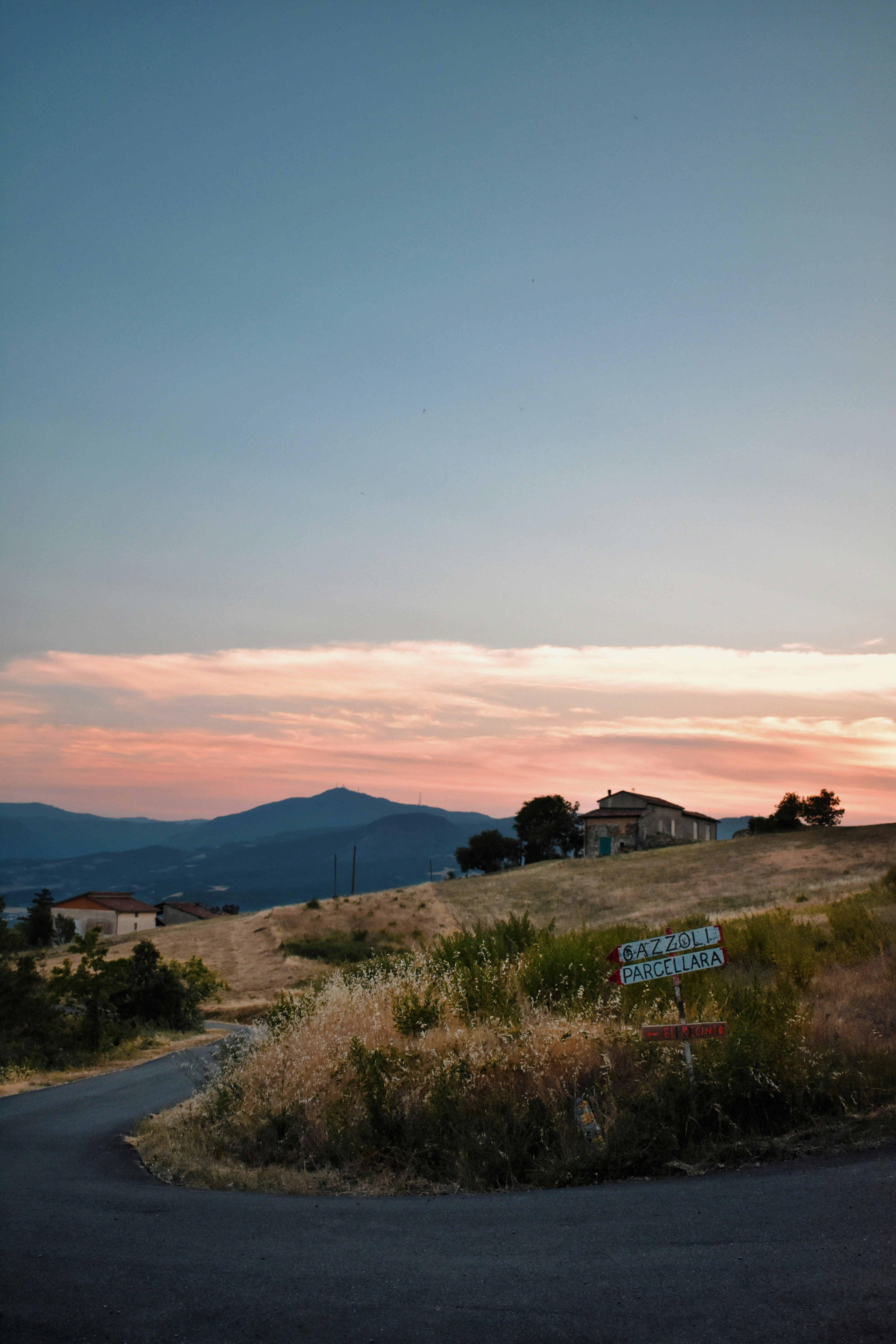Winding road leading through a rural landscape at sunset with distant mountains and a farmhouse.