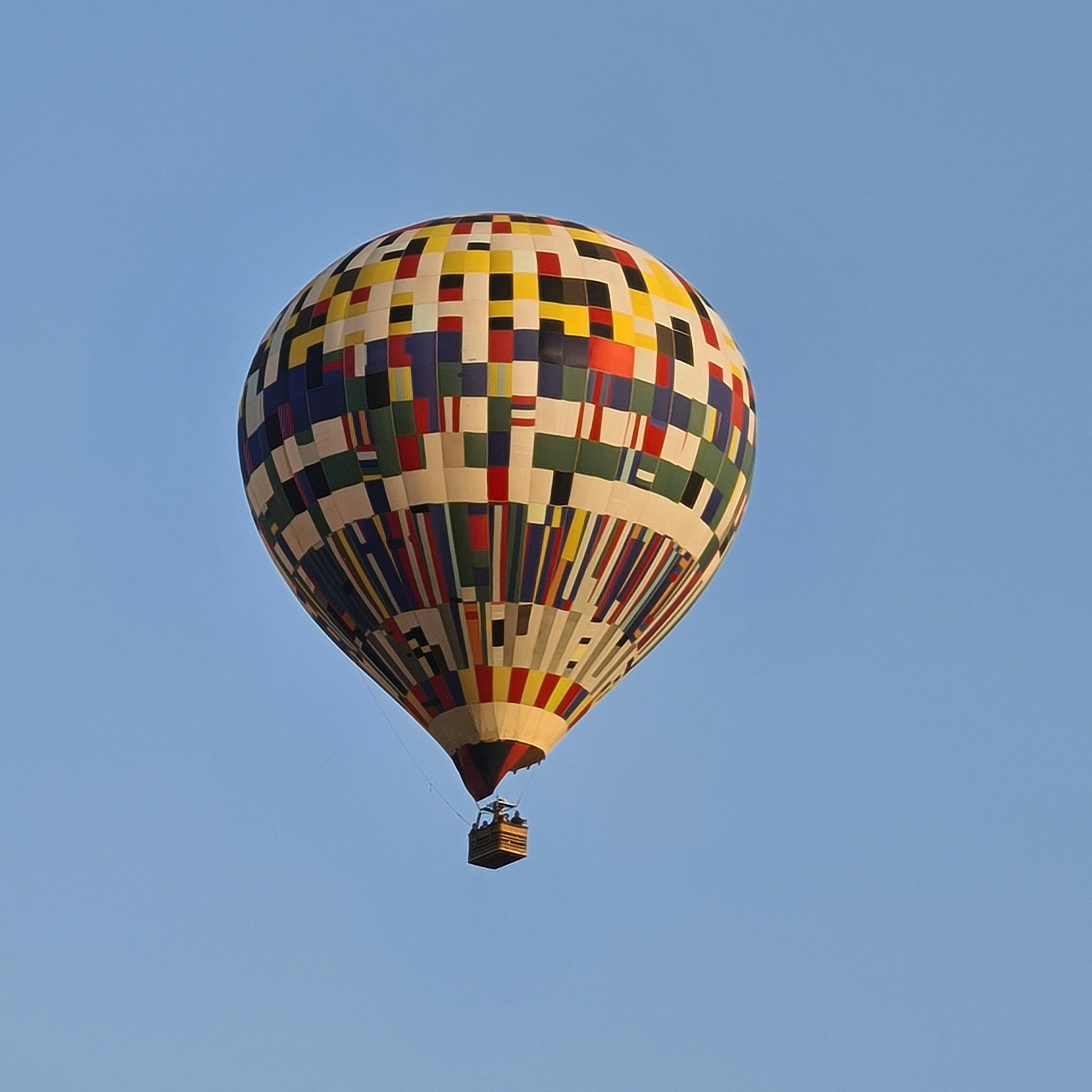 Colorful hot air balloon with a patchwork pattern rises against a clear blue sky, photograph.