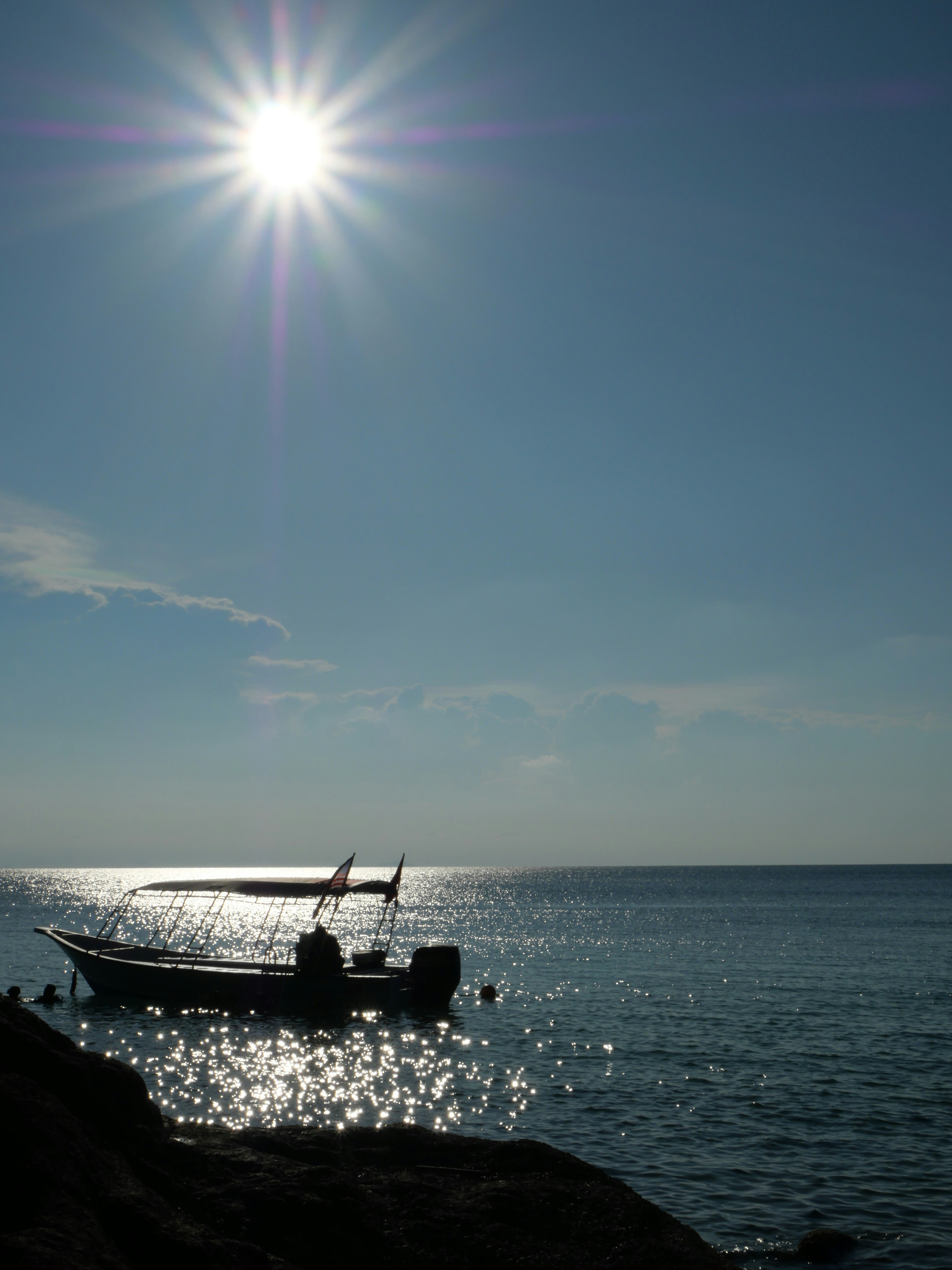 Photograph of a silhouette boat anchored near a rocky shore under a starburst sun on a calm sea.