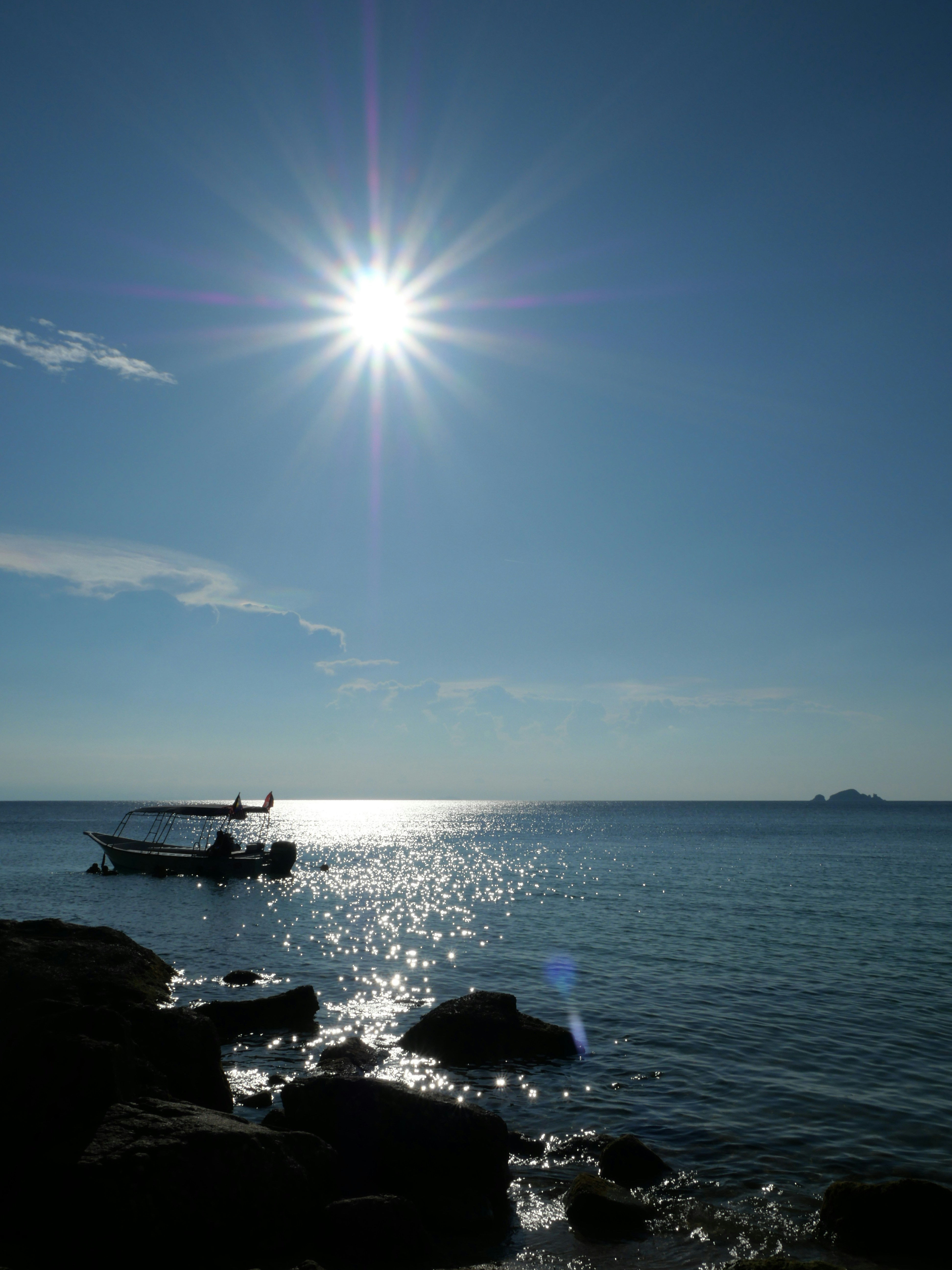 Fishing boat gliding across a shimmering sea under a radiant sun, with rocky shore framing the scene.