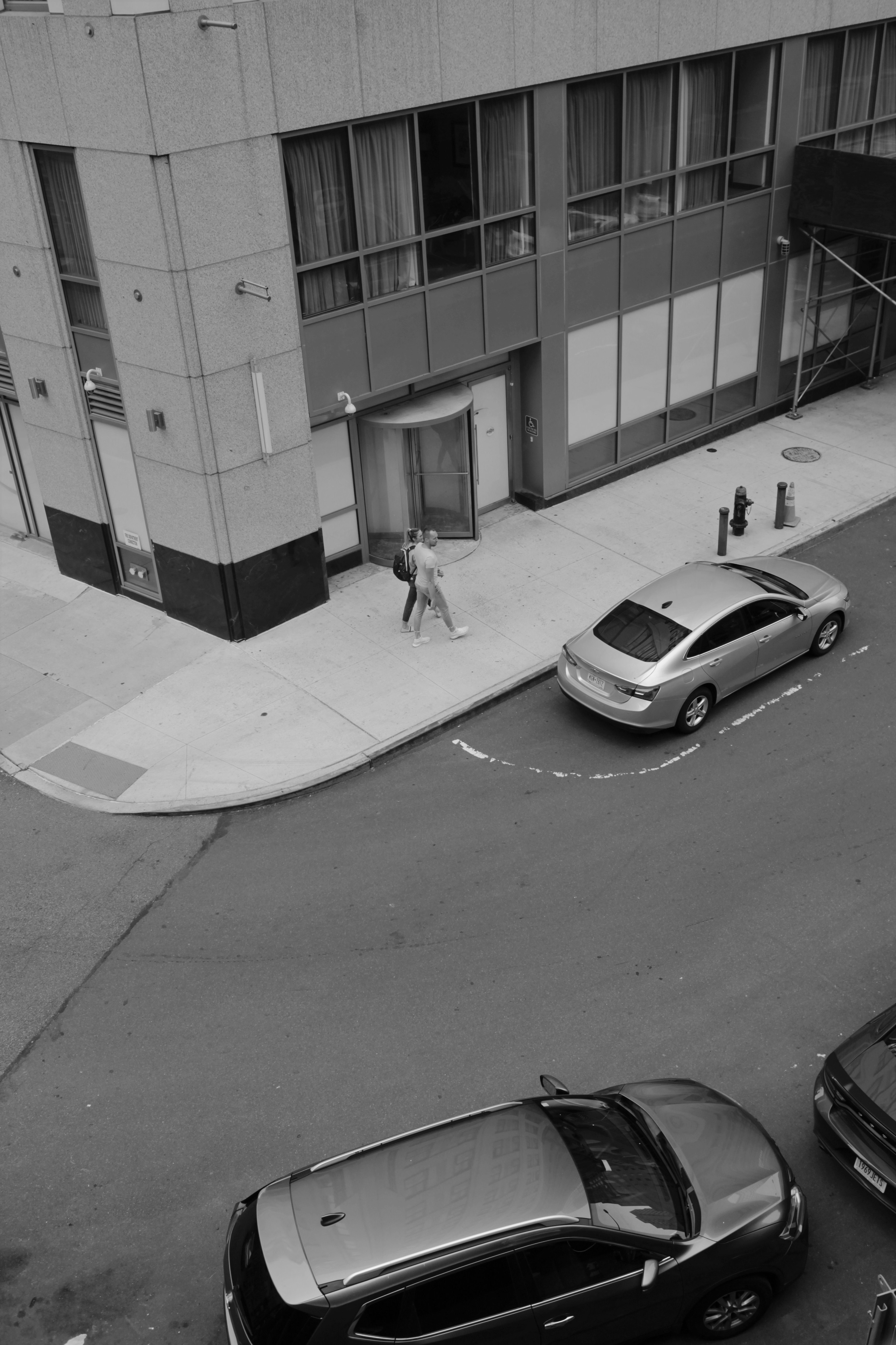 Monochrome street photograph showing two pedestrians near a modern building entrance as a silver sedan is parked along the curb.