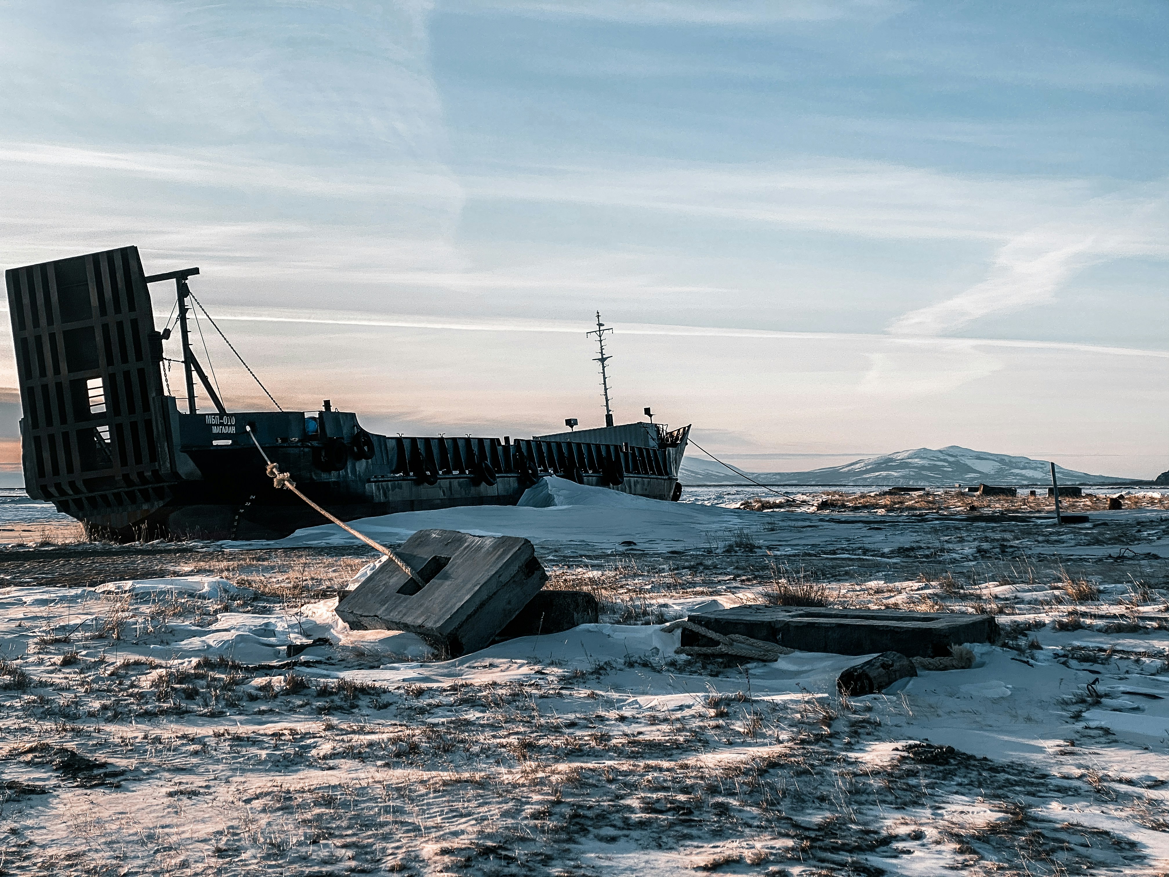 Abandoned shipwreck resting on a snowy landscape with distant mountains under a pale sky.