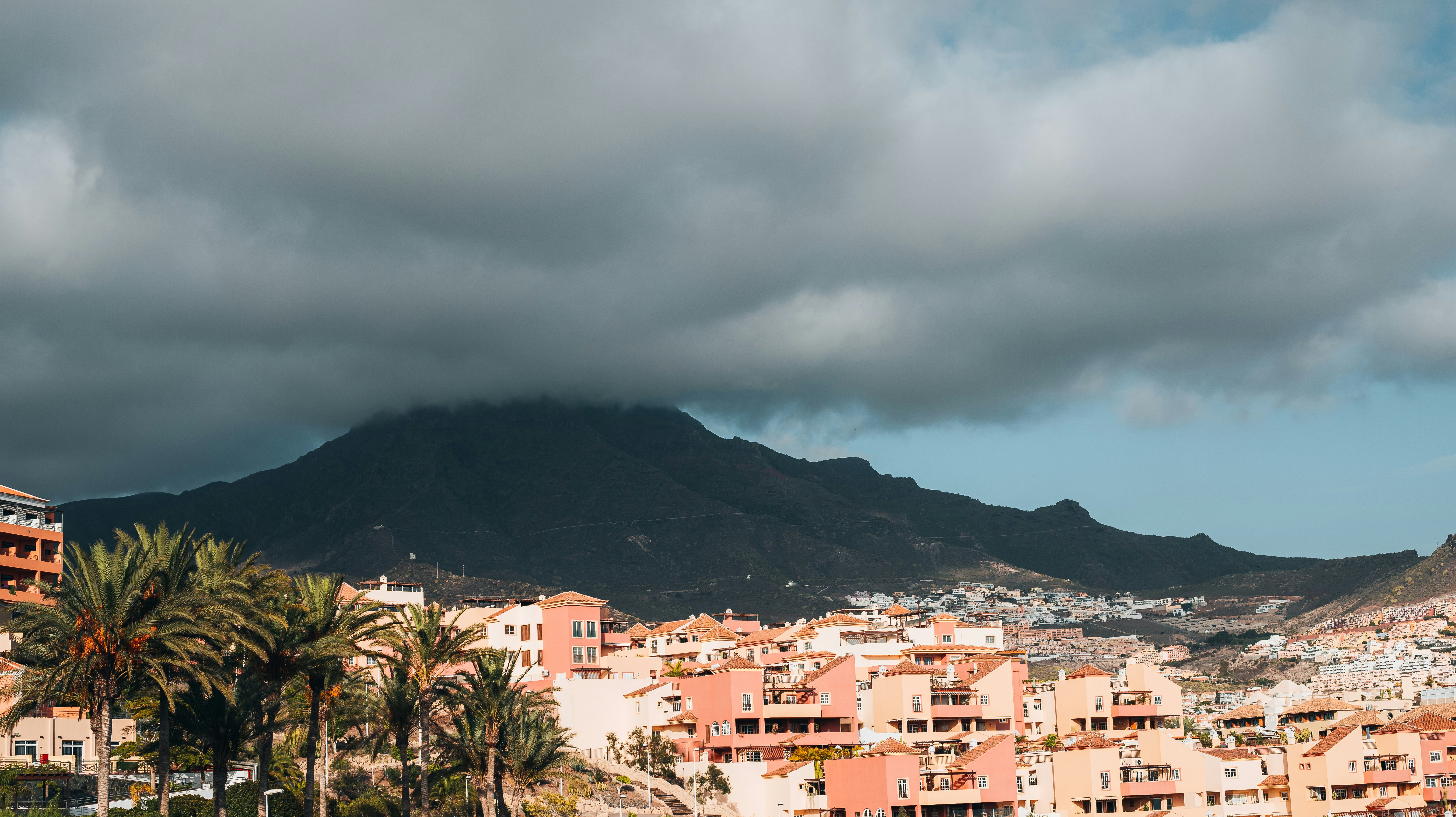 Cityscape with terracotta buildings, palm trees, and mountains under dramatic clouds.