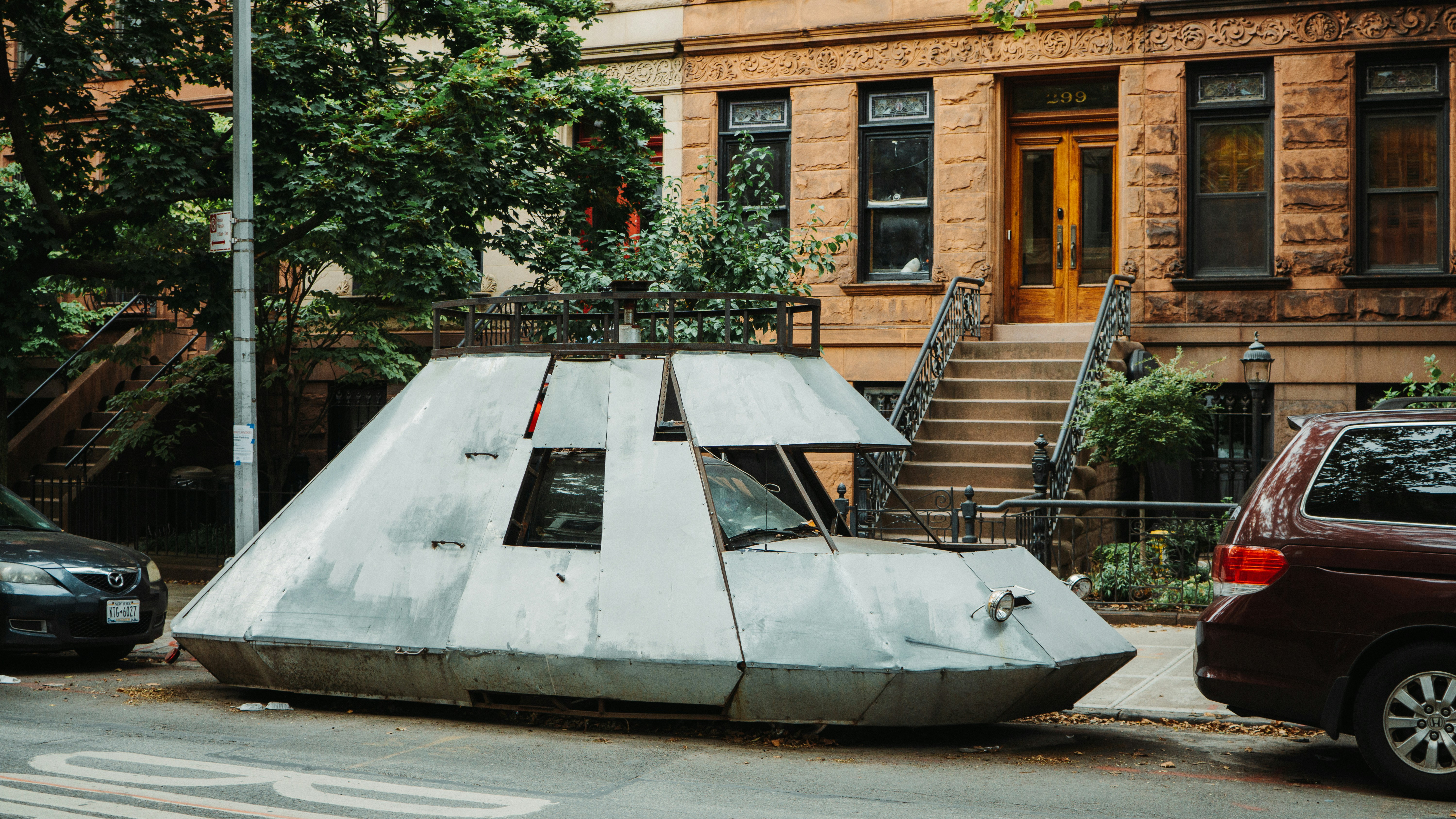 An unconventional, metallic vehicle resembling a futuristic craft parked on a city street, surrounded by classic brownstone architecture.
