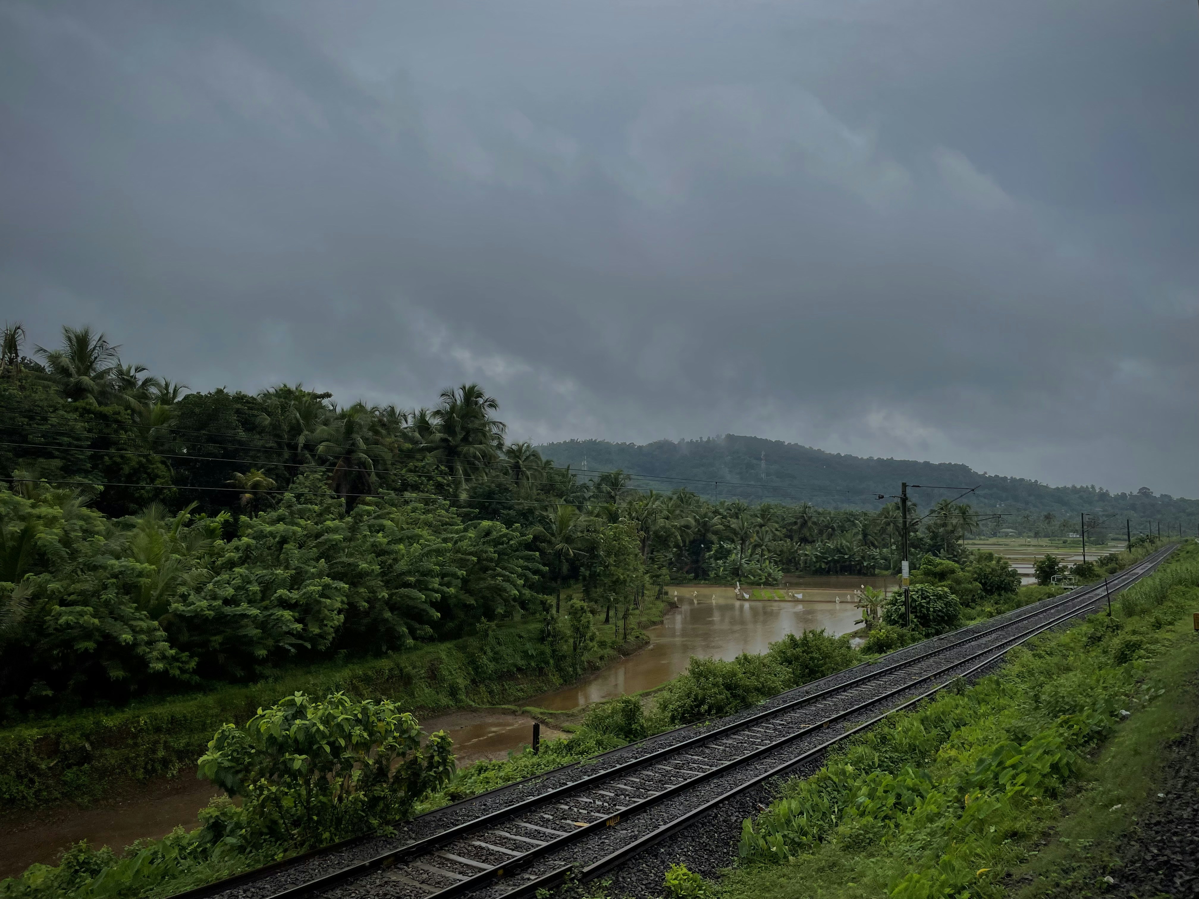 Lush greenery flanks a railway track beside a calm, rain-fed river under a moody sky. The scene evokes tranquility amidst the approaching storm.