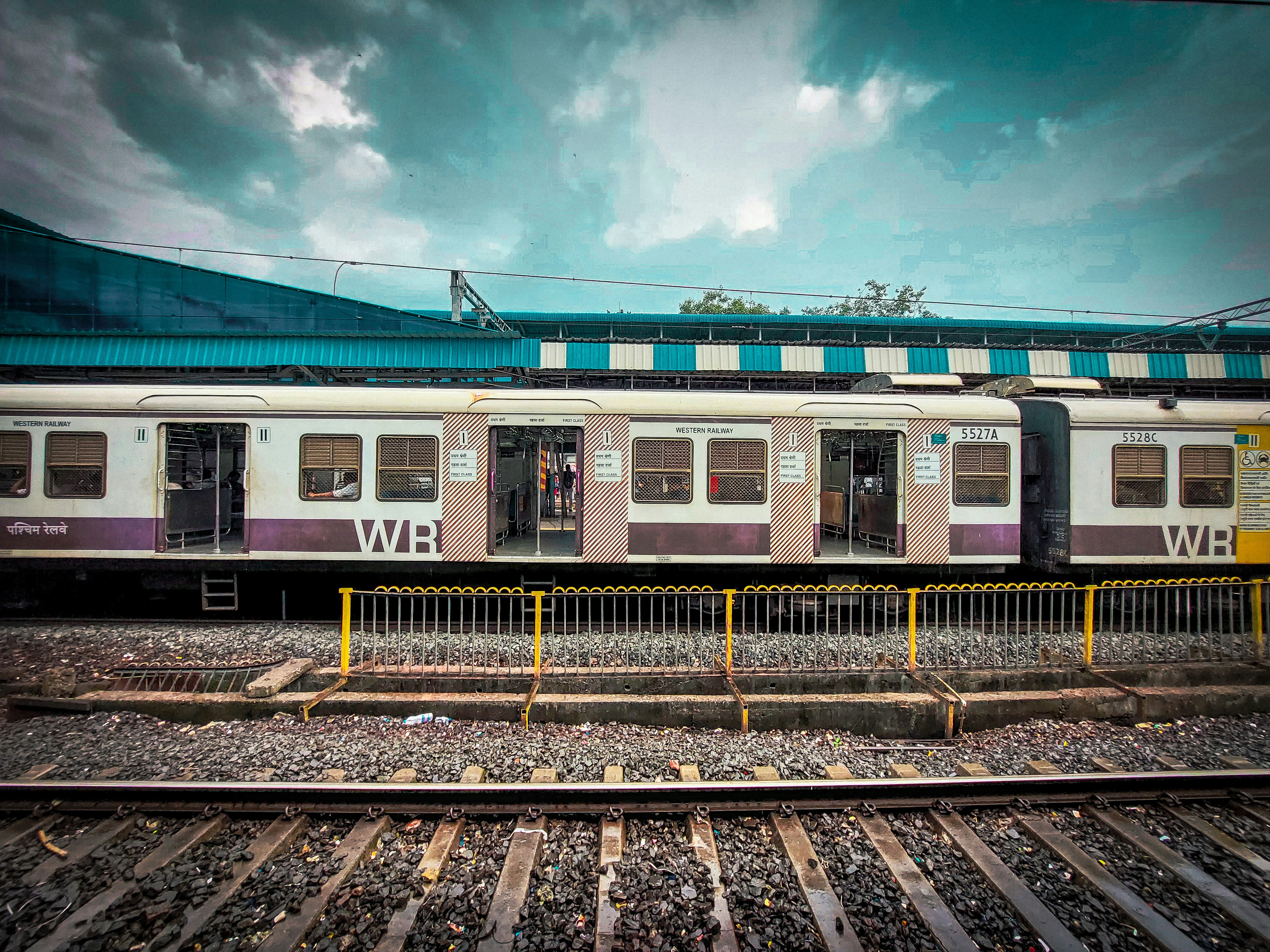 Train carriage with doors ajar sits on a platform beside yellow safety rails, with parallel tracks in the foreground. The sky overhead is a vivid turquoise, lending a cinematic transit vibe.