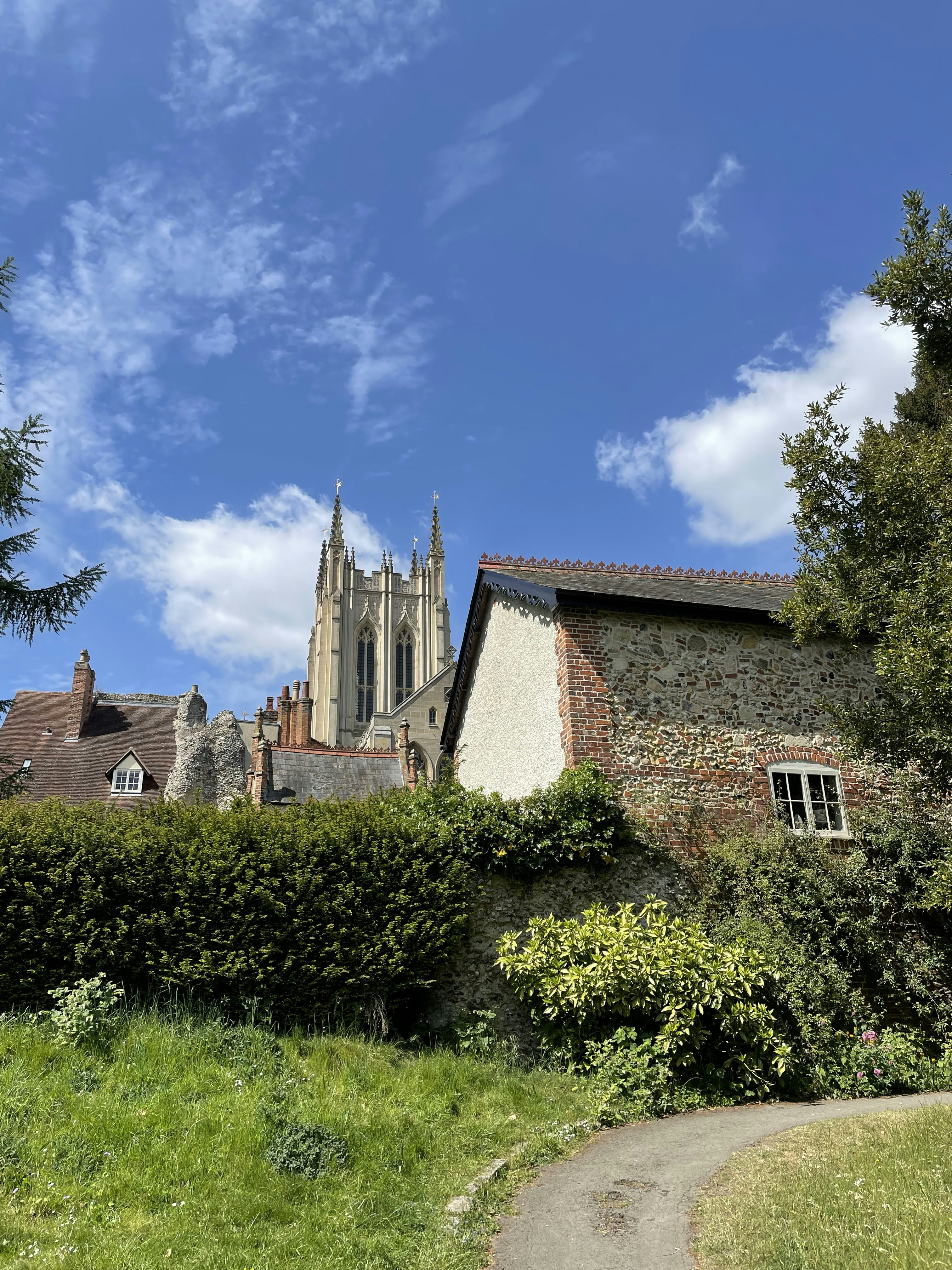 Historic church tower rising above lush greenery and rustic buildings under a bright blue sky.