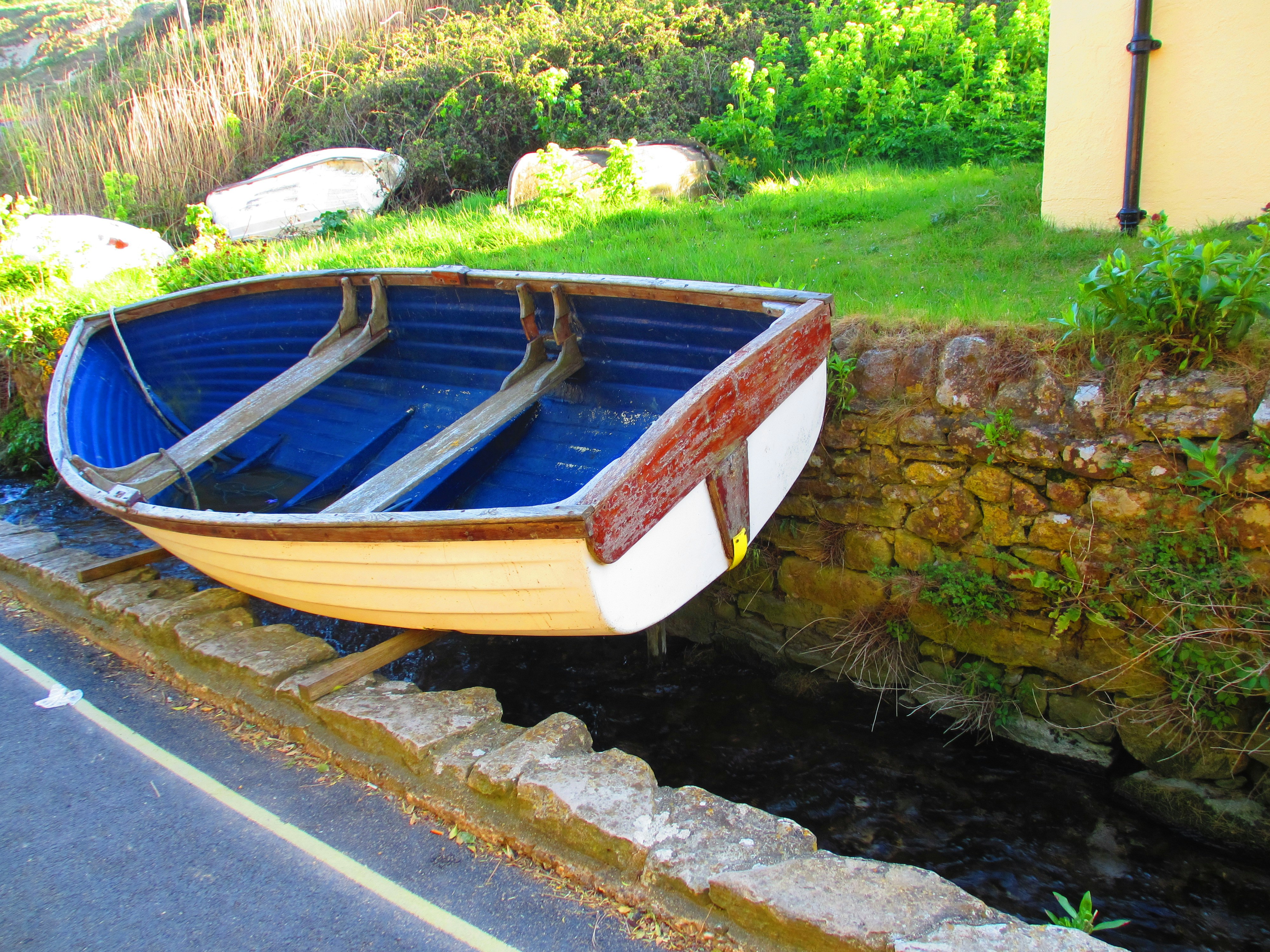 An old wooden boat rests on a stone ledge beside a gently flowing stream, surrounded by lush greenery.