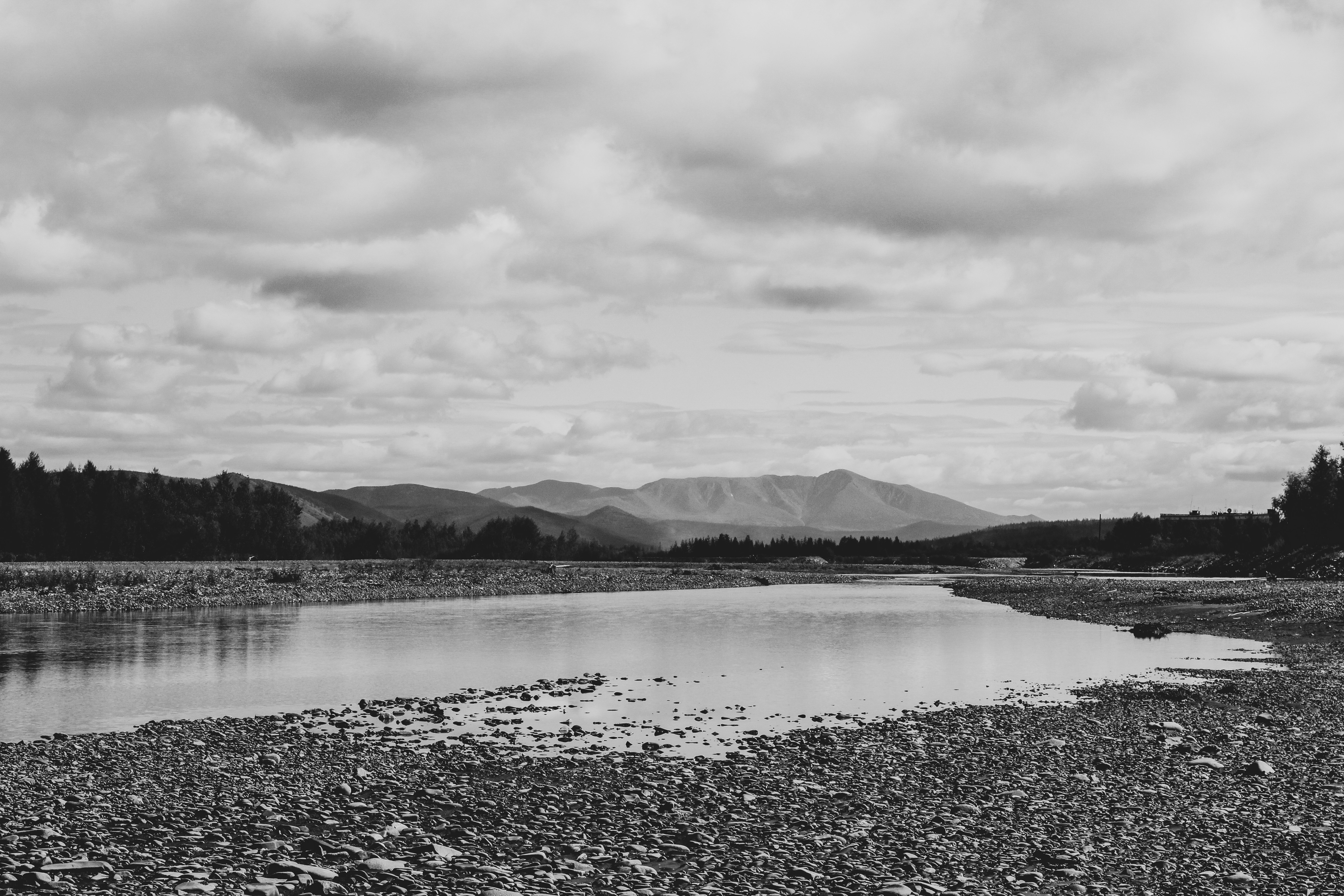 Black and white landscape of a tranquil river with distant mountains under a cloudy sky.