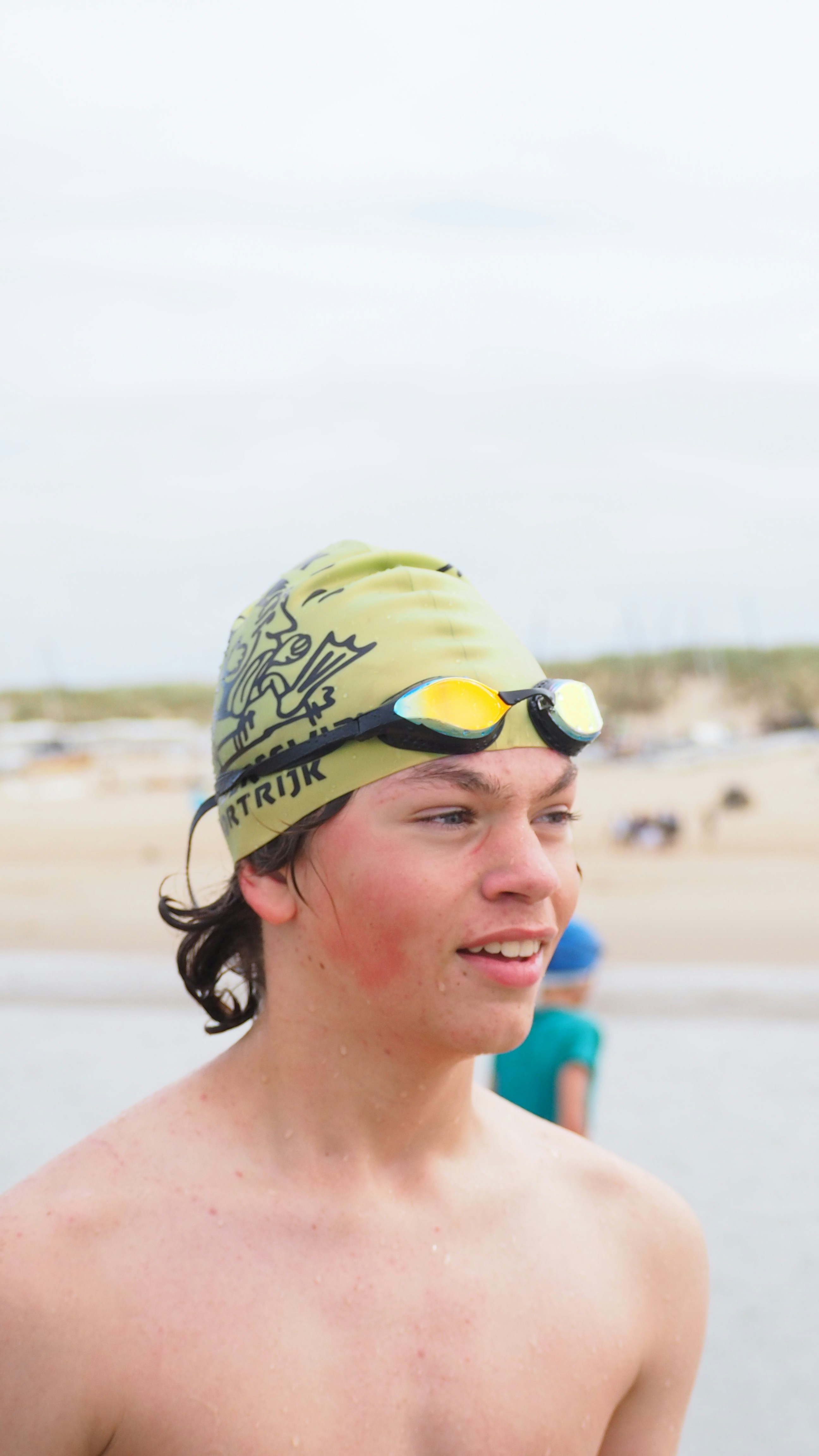 Young swimmer with goggles looks out over a sandy beach under an overcast sky.