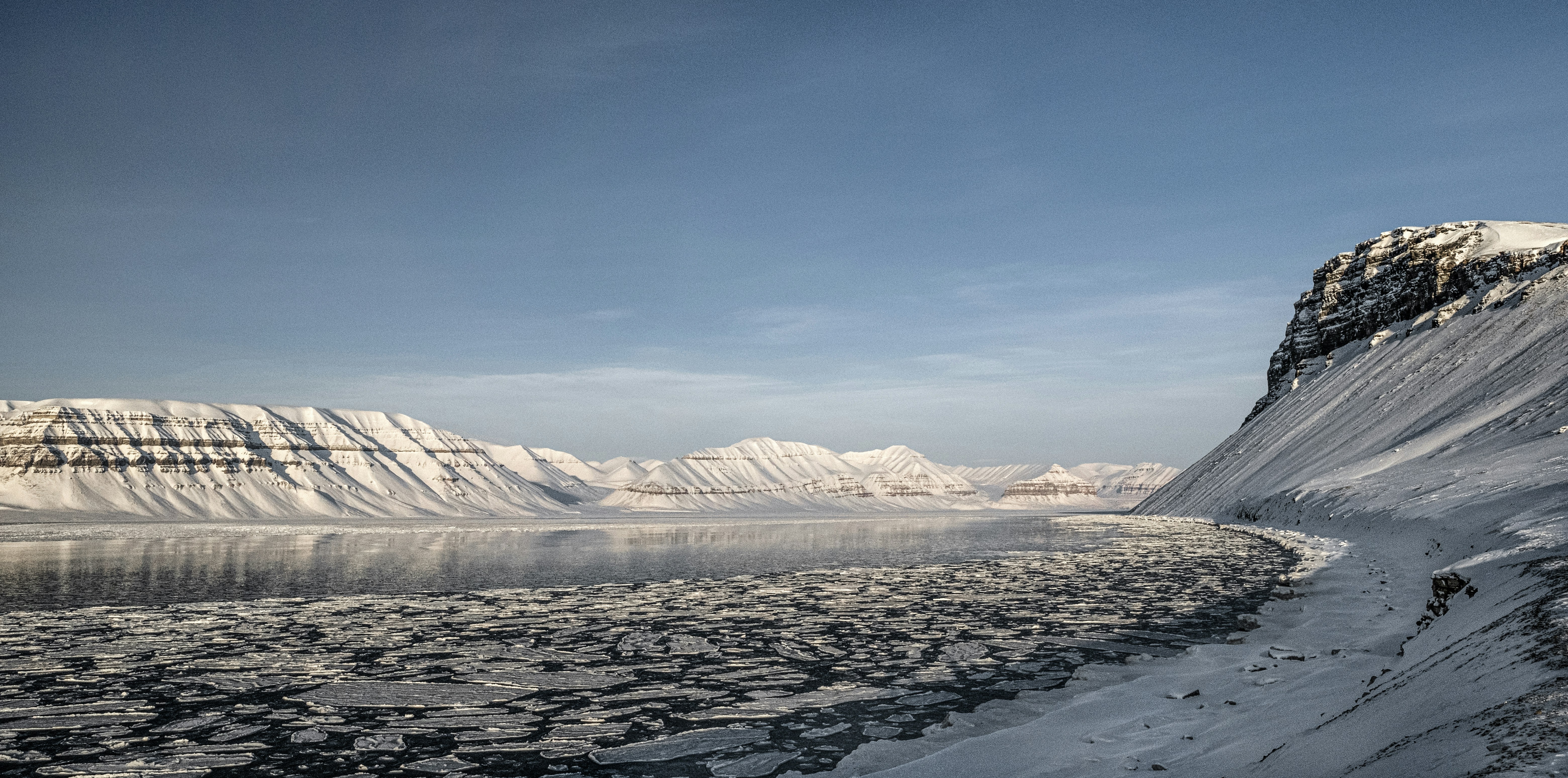 Ice floes drift along the fjords under a clear blue sky, bordered by snow-capped mountains.