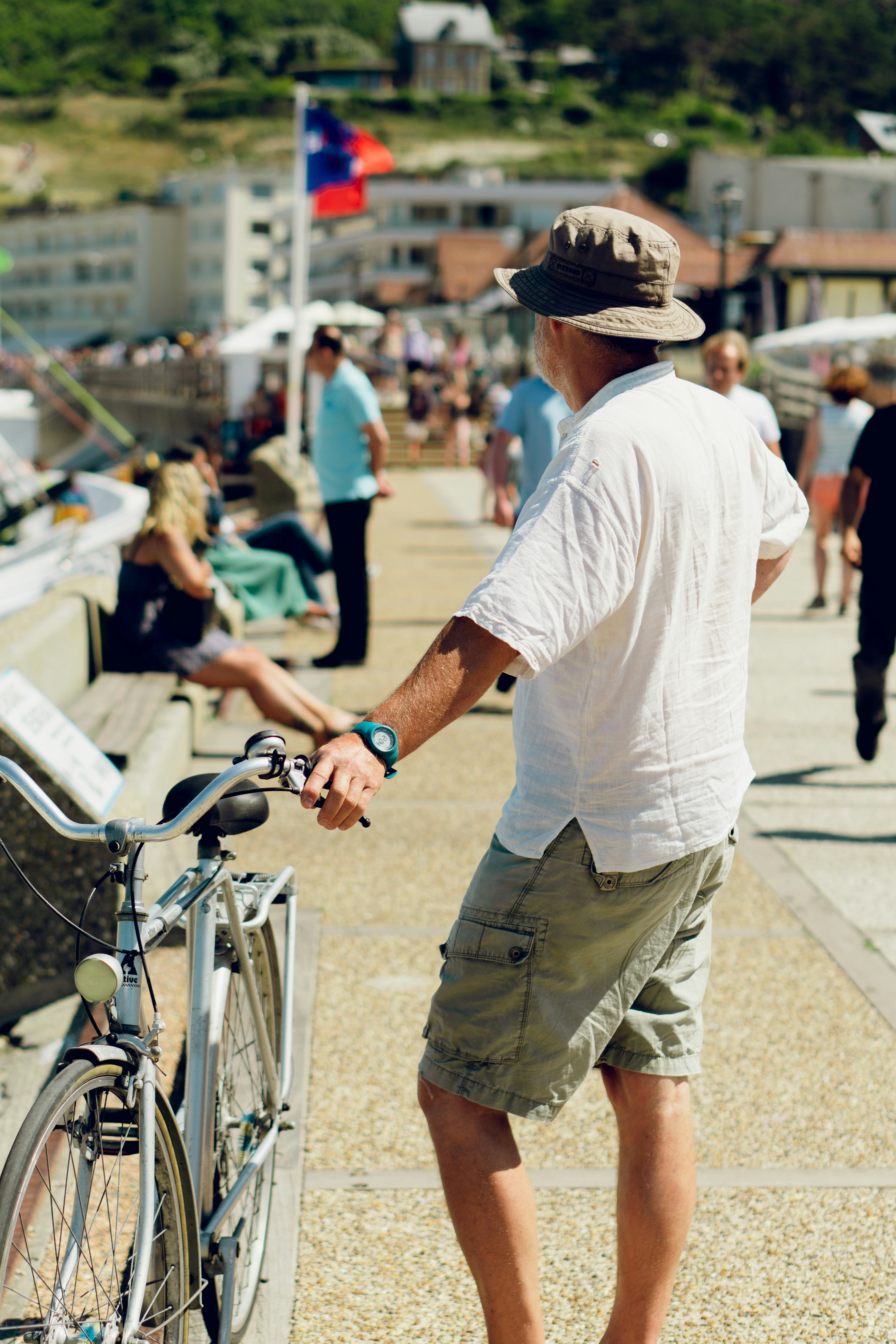 Man in a sun hat holding a bicycle, walking along a bustling promenade with people enjoying the sunny day. Colorful flags flutter in the background.