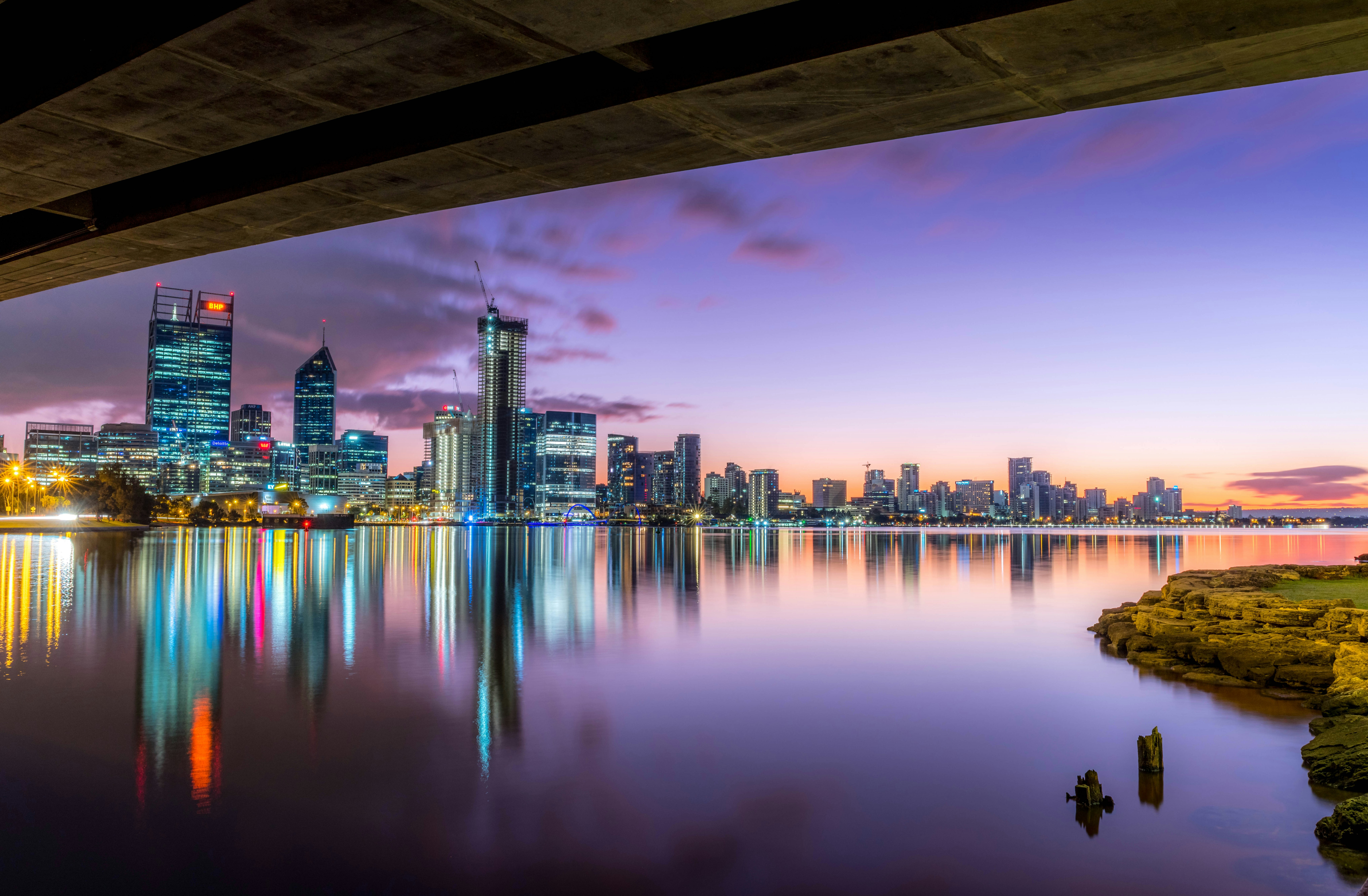 City skyline illuminated at twilight, reflecting on calm waters beneath a bridge. Vibrant colors create a serene atmosphere.