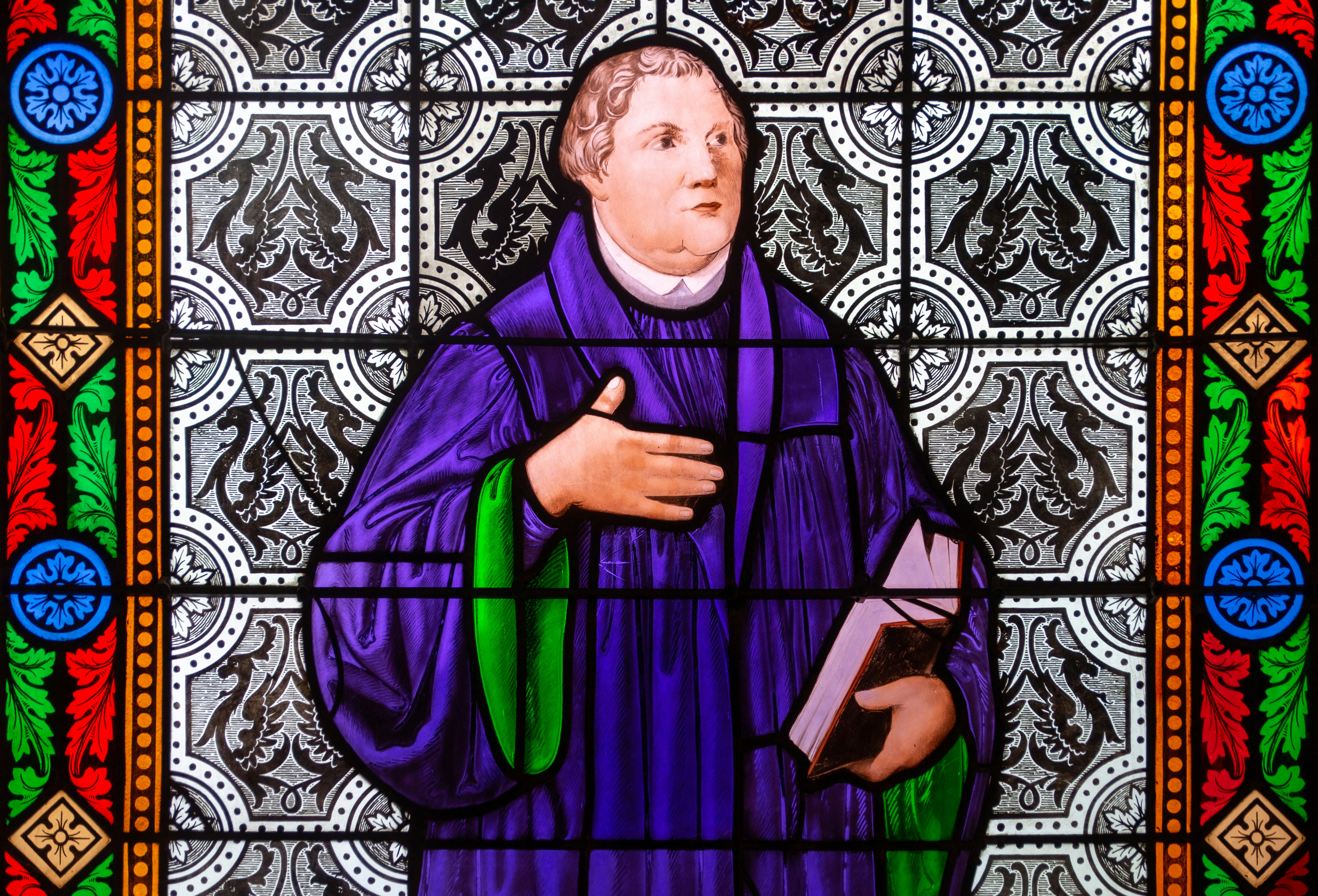Stained glass window depicting a clergyman in a purple robe holding a book, surrounded by intricate geometric patterns.
