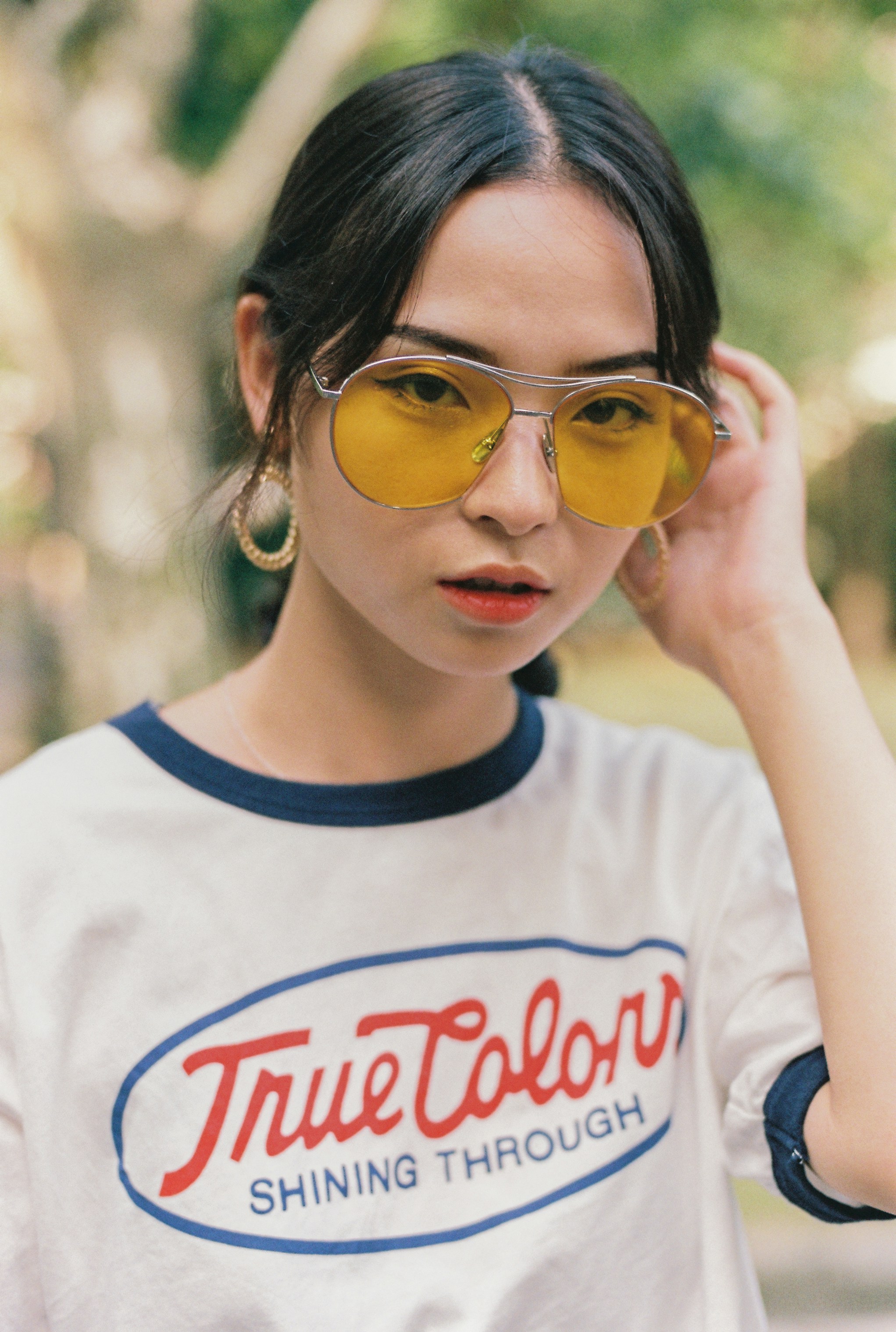 Young woman with yellow sunglasses and a retro 'True Colors' shirt poses against a blurred green backdrop.