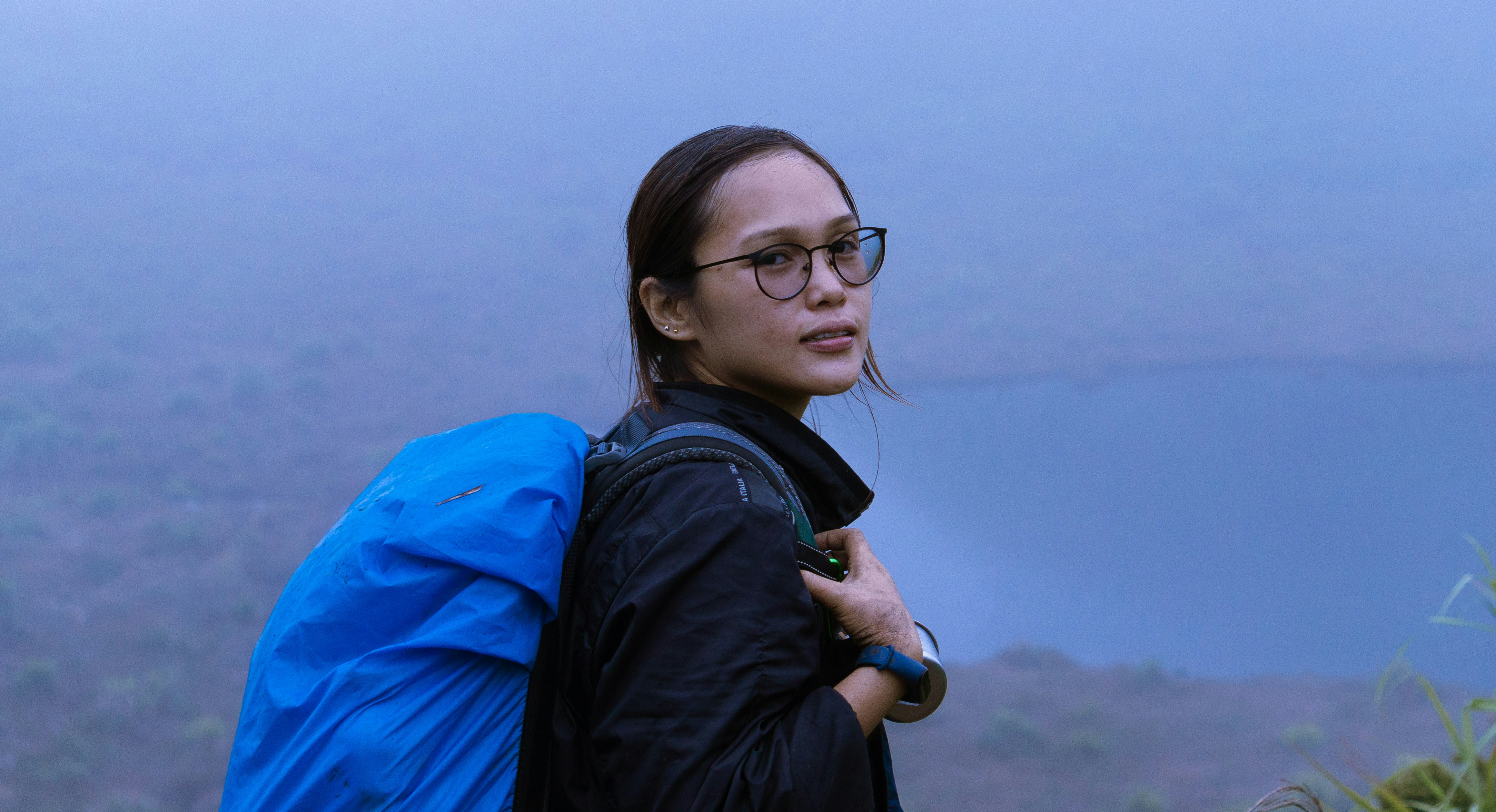 Portrait of a hiker with a blue backpack standing in a misty blue-toned landscape.