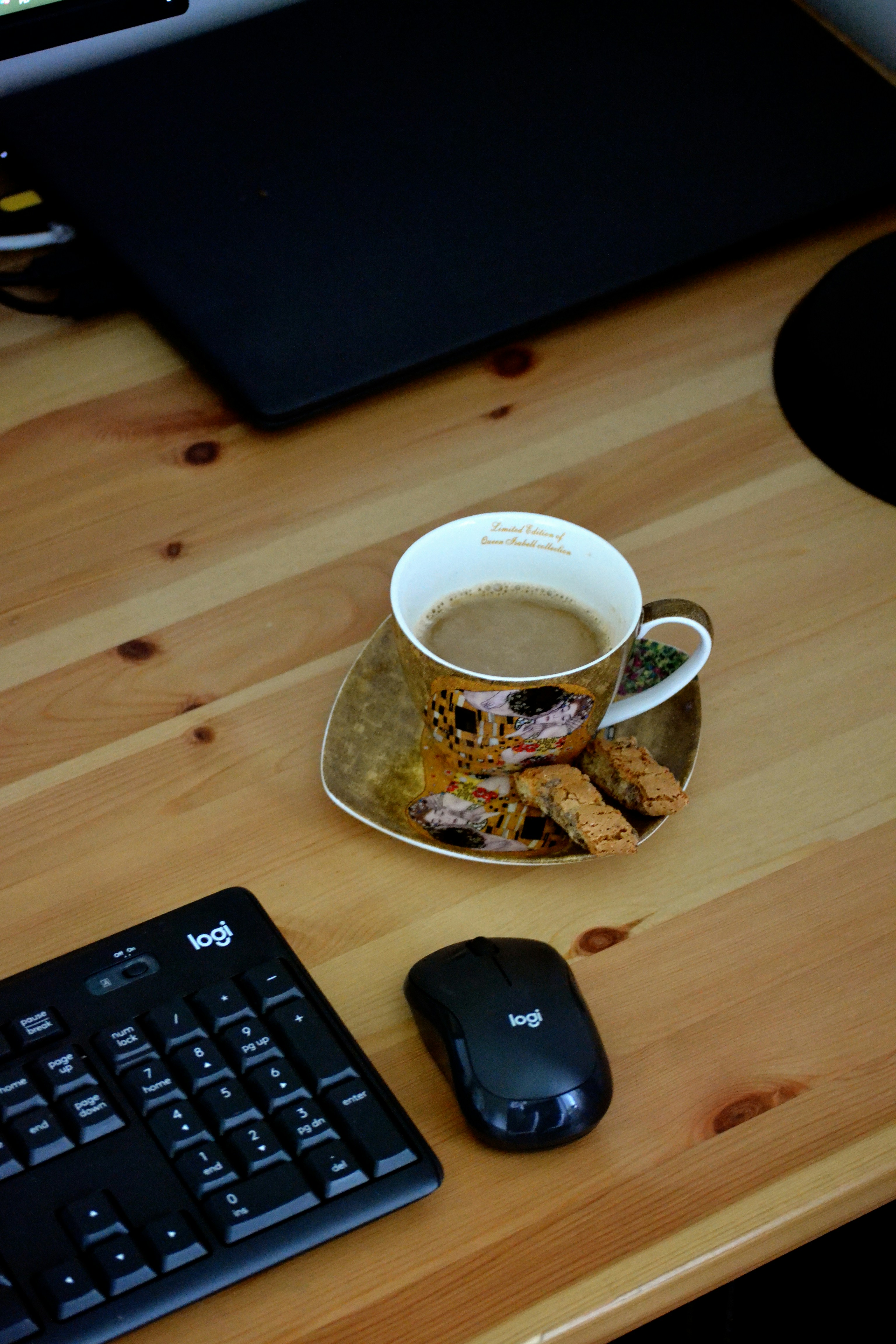 Coffee cup with a decorative saucer and cookies rests on a wooden desk beside a keyboard and mouse.