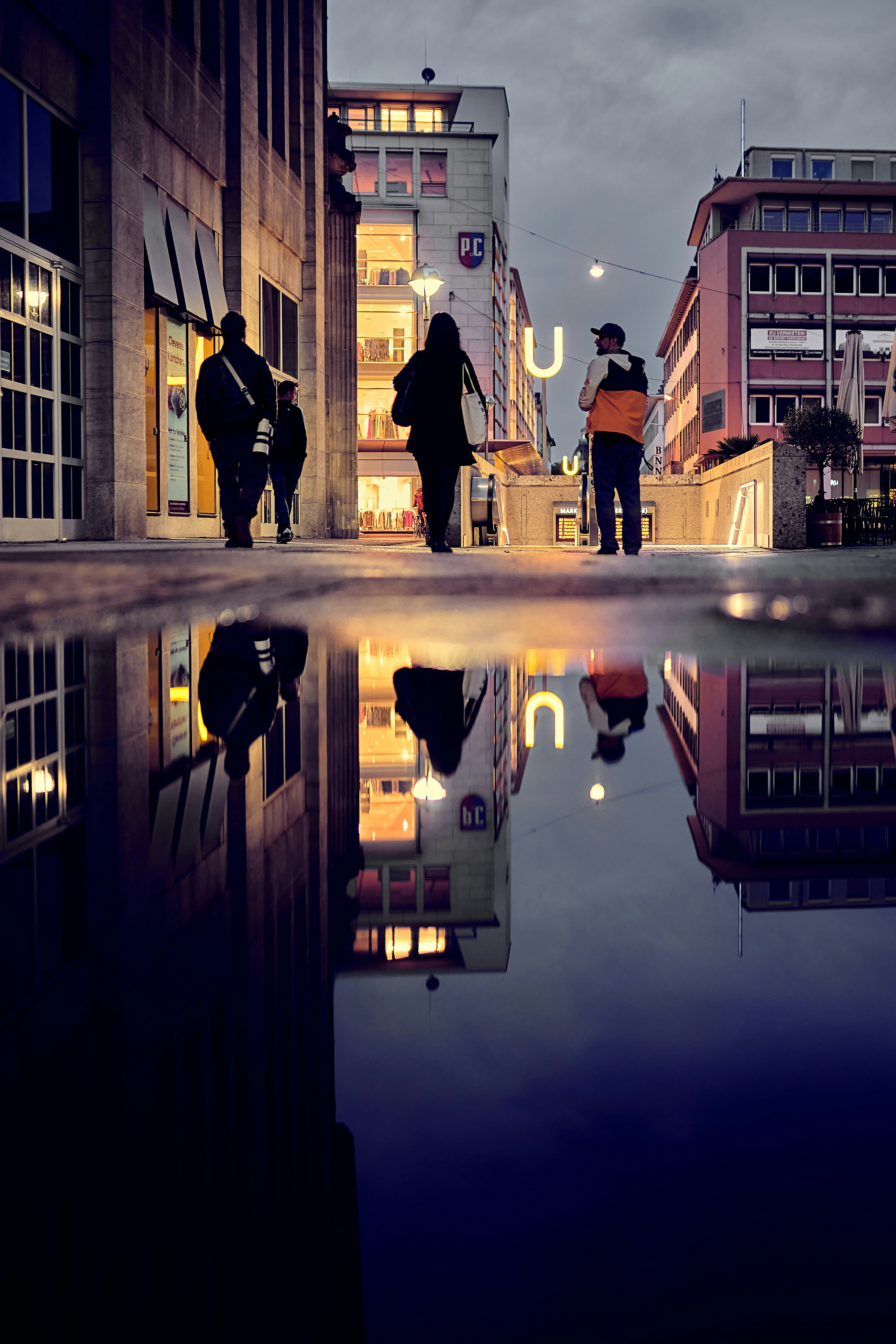Silhouetted figures and illuminated buildings reflected in a puddle on a city street at dusk.