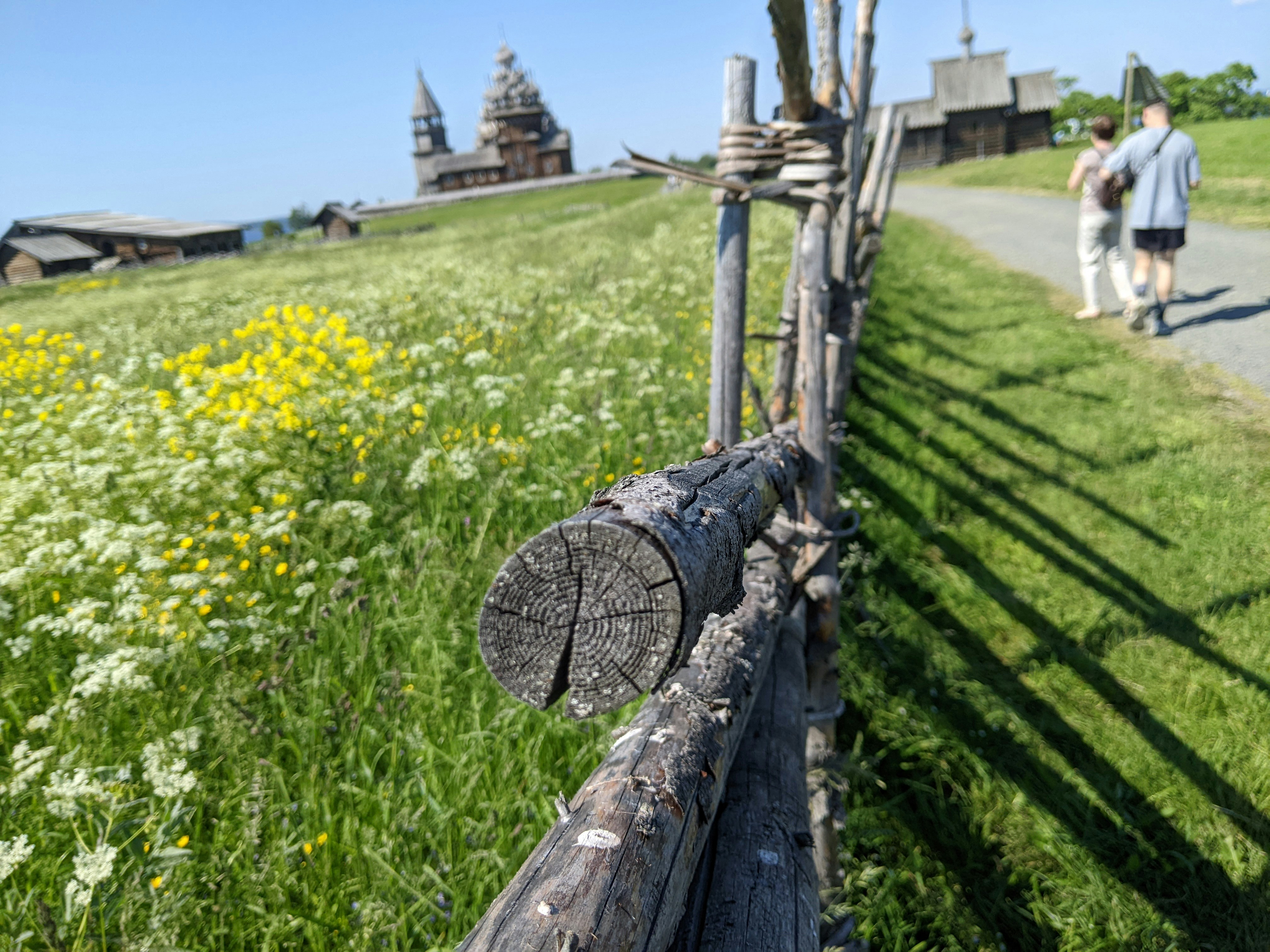 Weathered wooden fence in the foreground, leading to a vibrant meadow with wildflowers and traditional wooden structures in the distance.