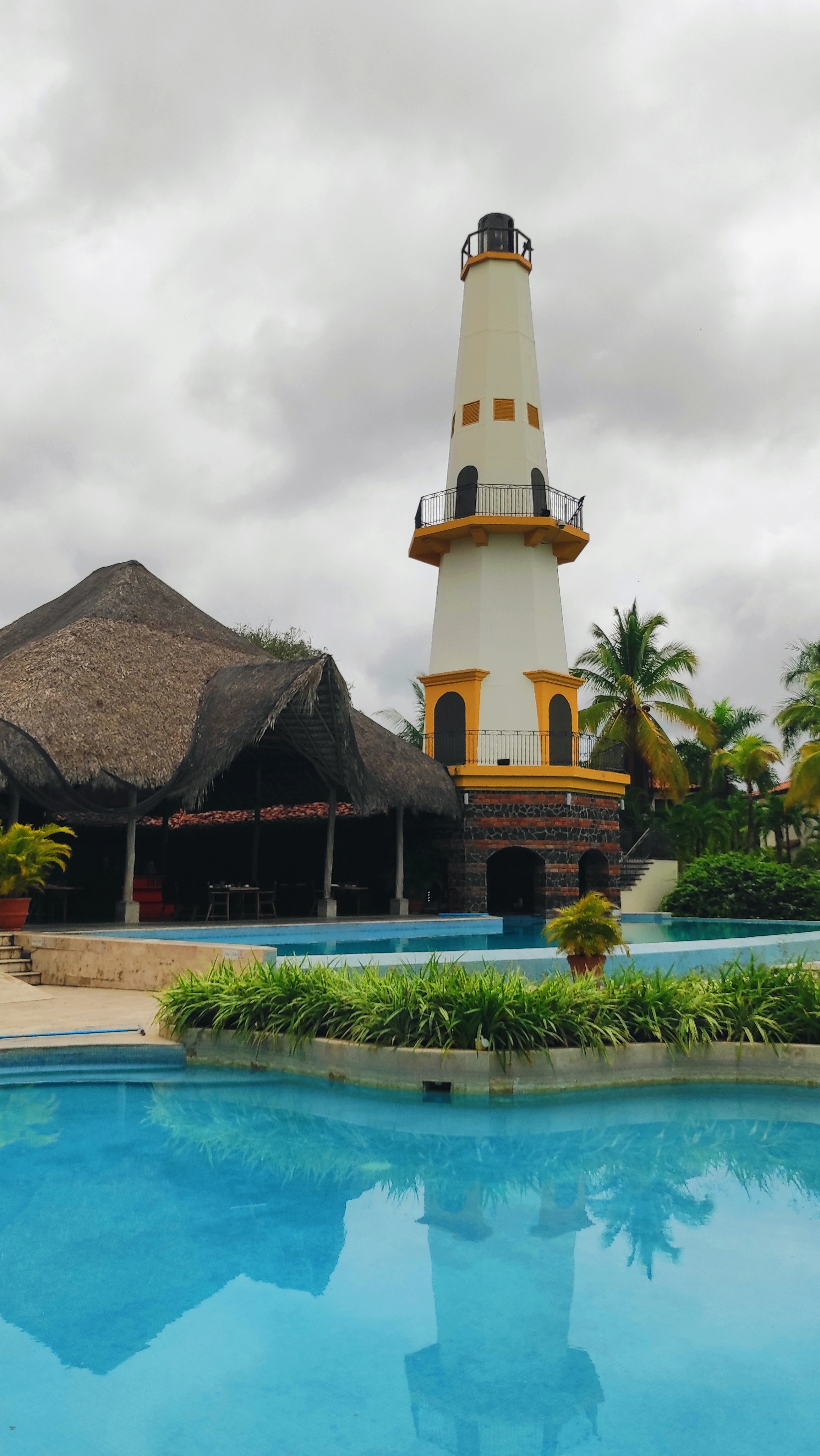 A lighthouse stands tall beside a tranquil pool, surrounded by lush greenery and palm trees, under a cloudy sky.