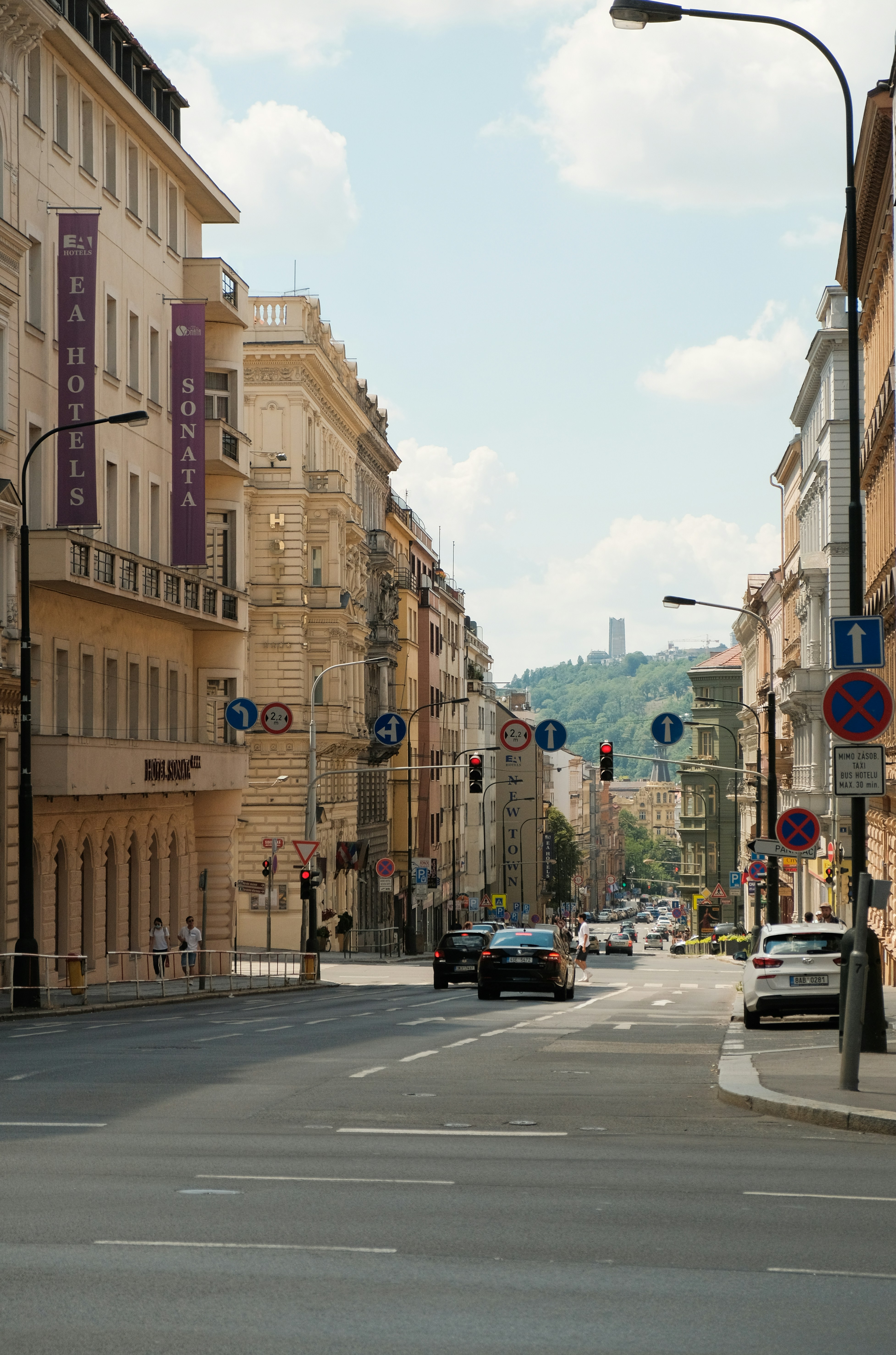 Busy street scene showcasing a blend of historic and modern architecture with traffic signals and pedestrians. A distant hill with a tower adds depth to the urban landscape.
