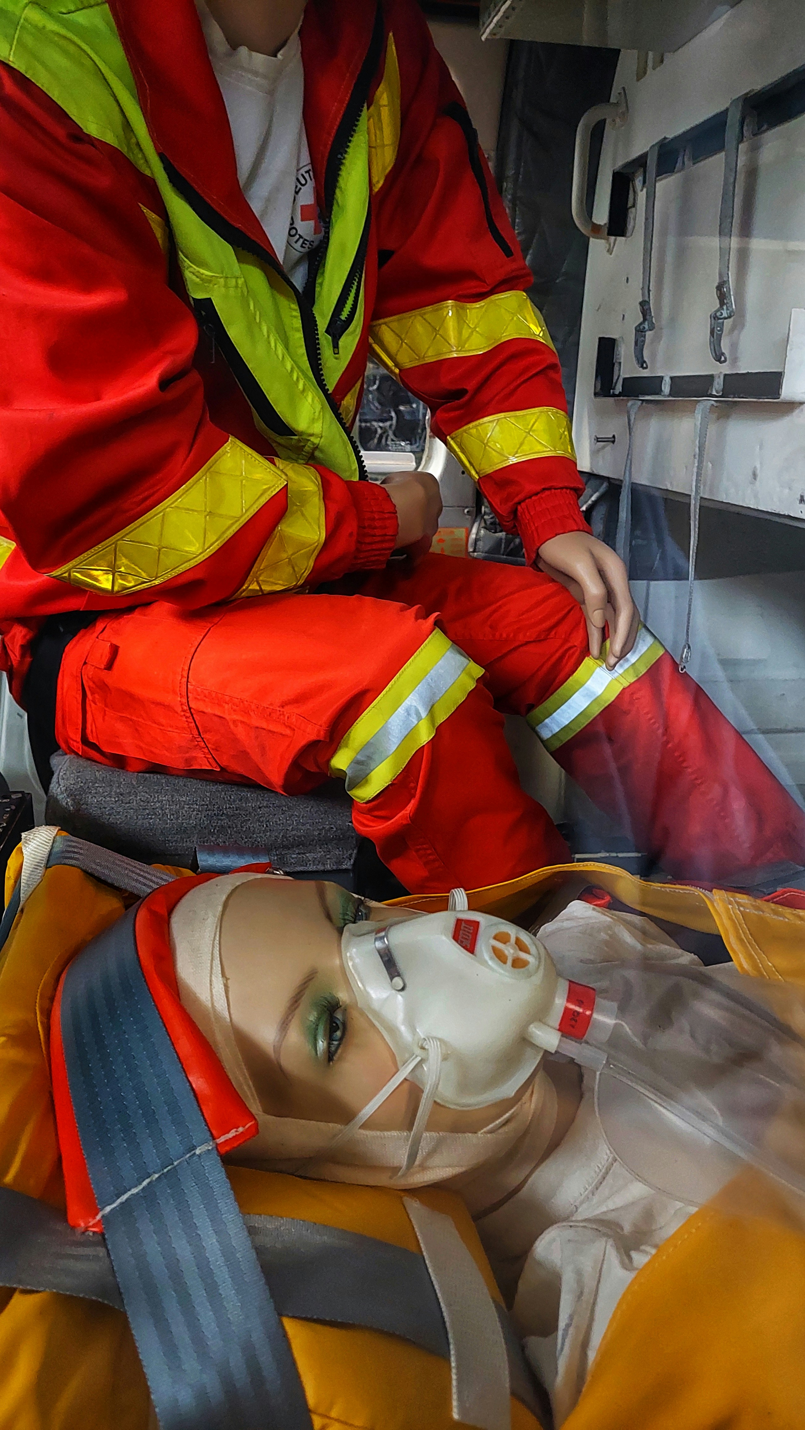 A rescue worker in bright protective gear sits beside a medical mannequin wearing a mask, simulating an emergency response scenario.