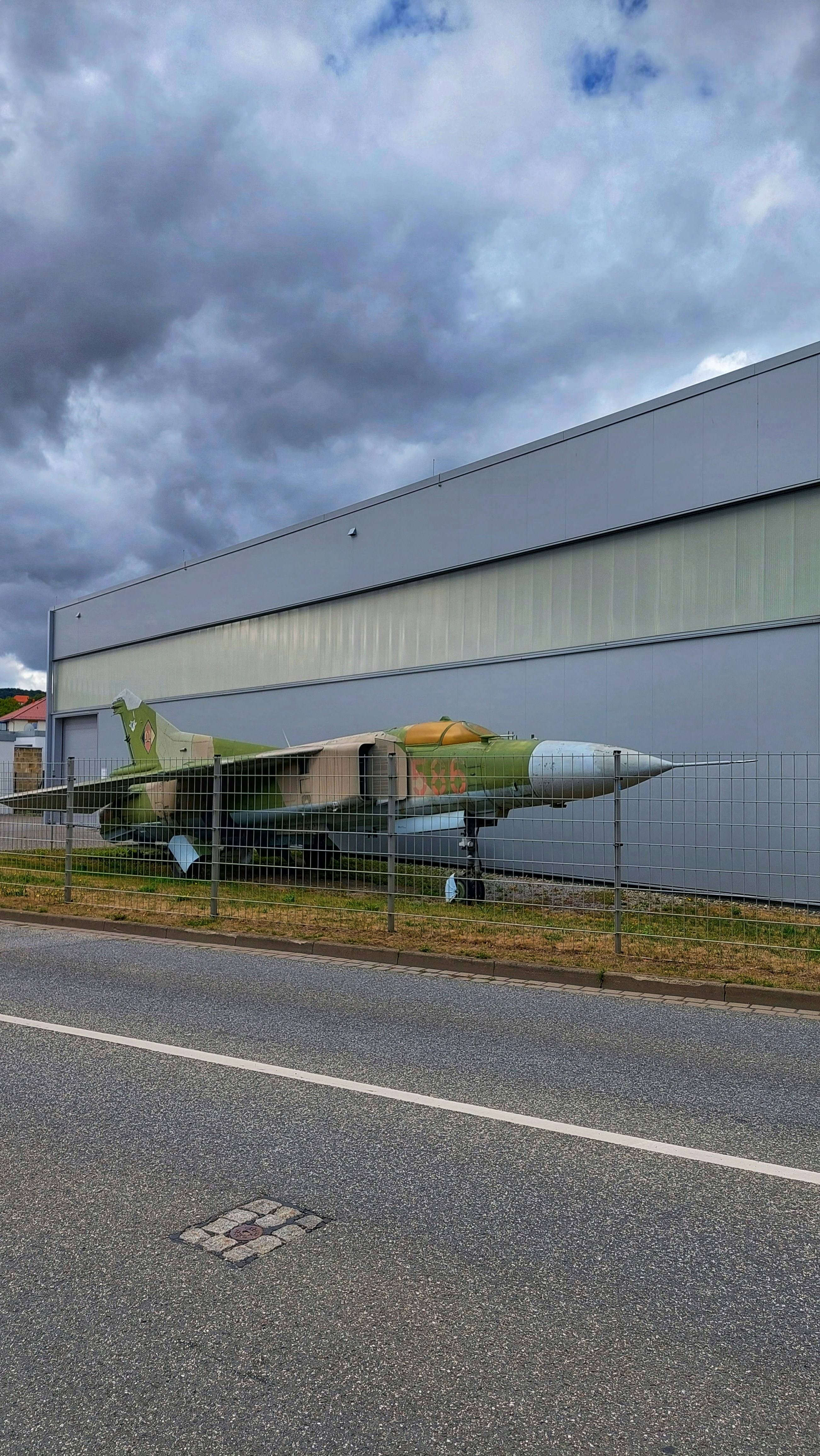 Camouflaged Fighter Jet Resting Beside a Fenced Hangar Under Moody Skies