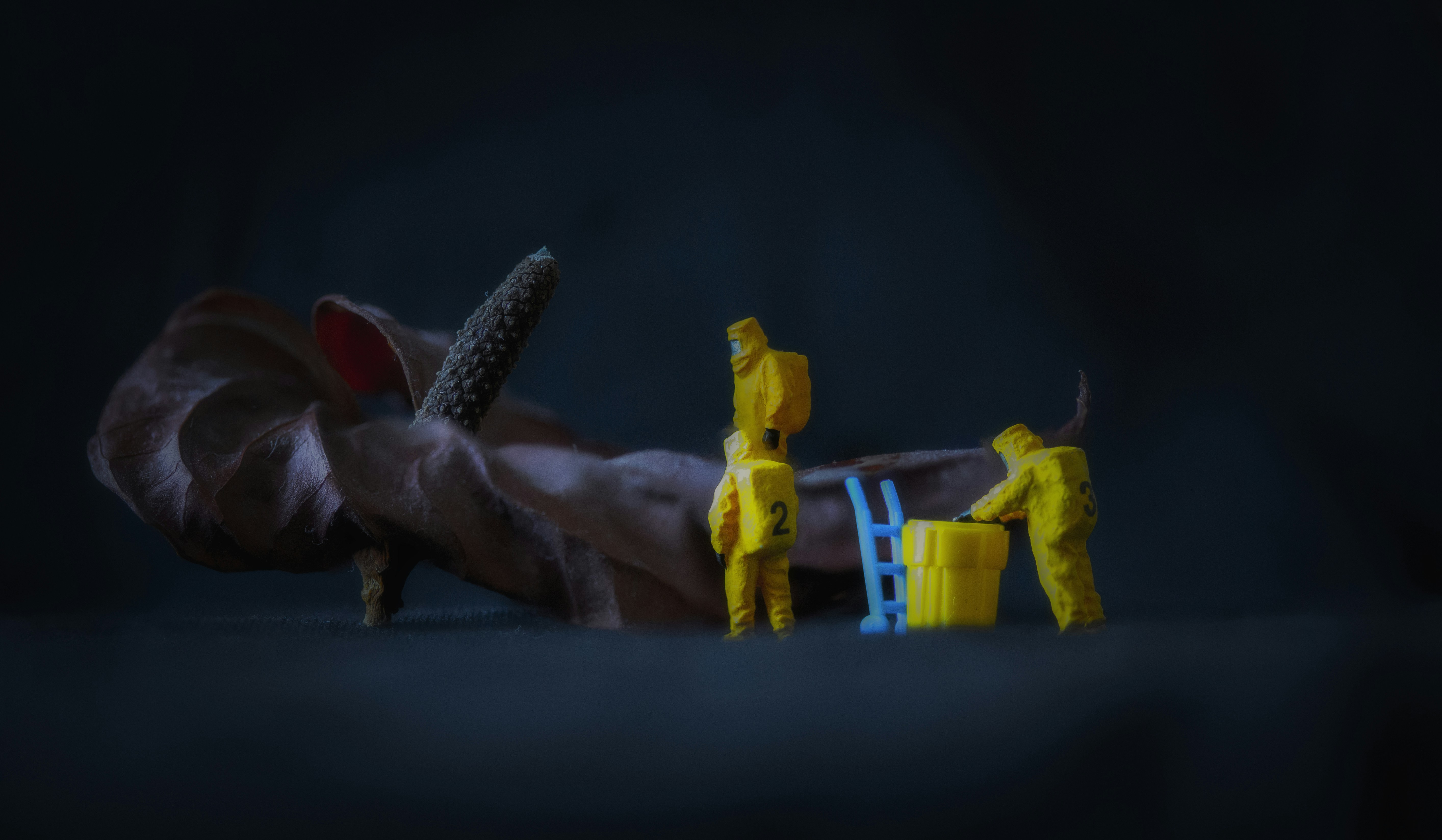 Two miniature hazmat figures interact with a vibrant flower petal and a small bucket, creating a whimsical scene of environmental care.