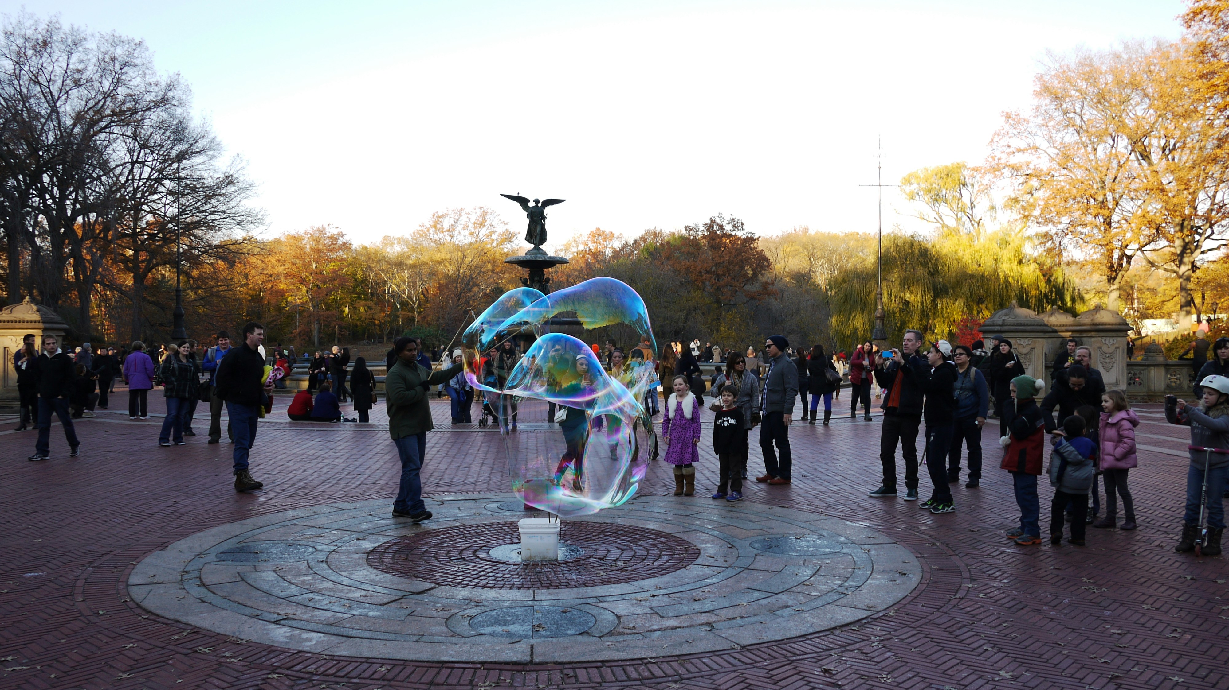 A large soap bubble floats above a crowd gathered around Bethesda Fountain in Central Park, capturing the joy of a vibrant autumn day.