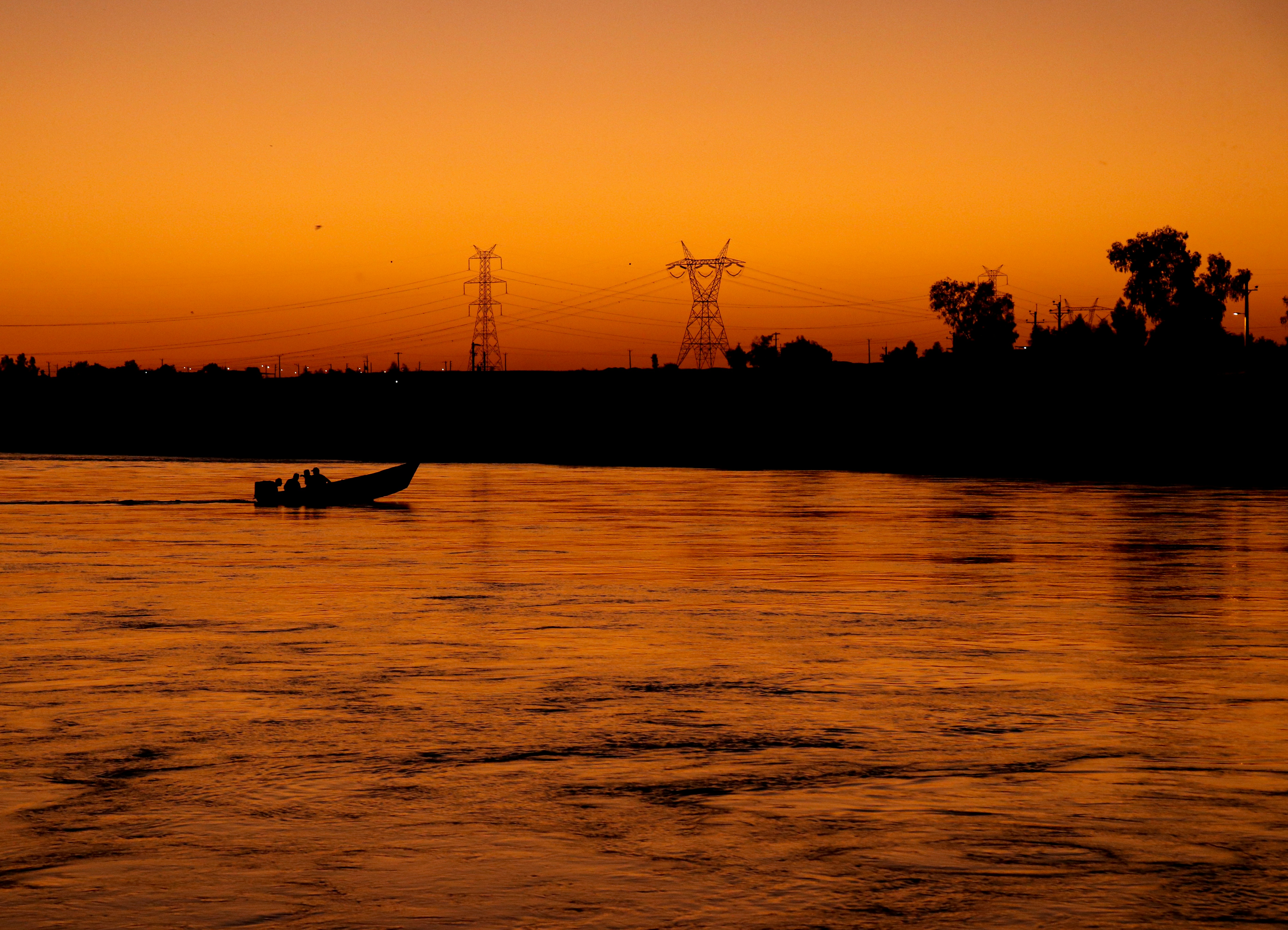 Silhouetted boat gliding on a river against a vibrant orange sunset with distant power lines.