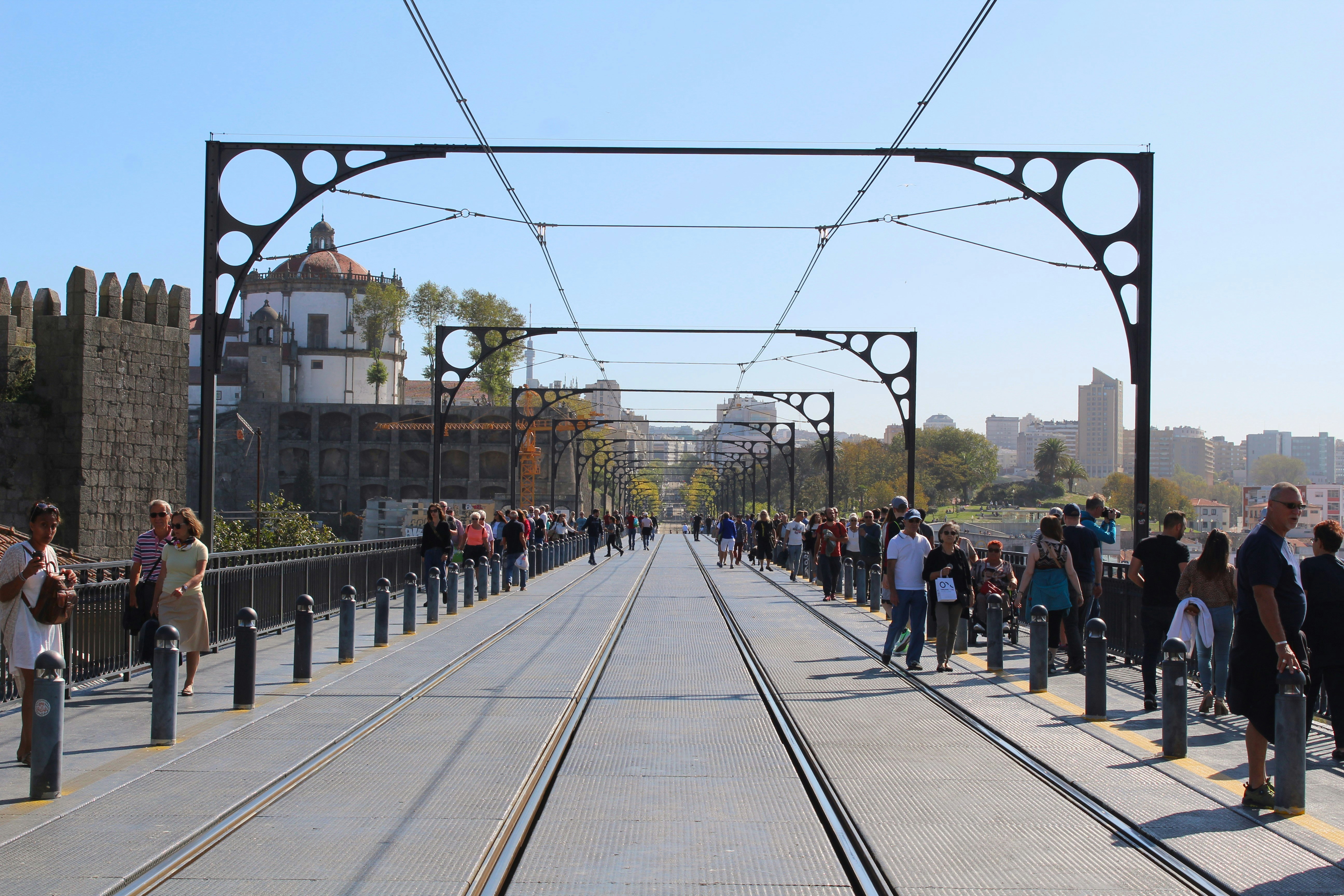 People walking on a wide bridge with intricate metalwork under a clear blue sky.