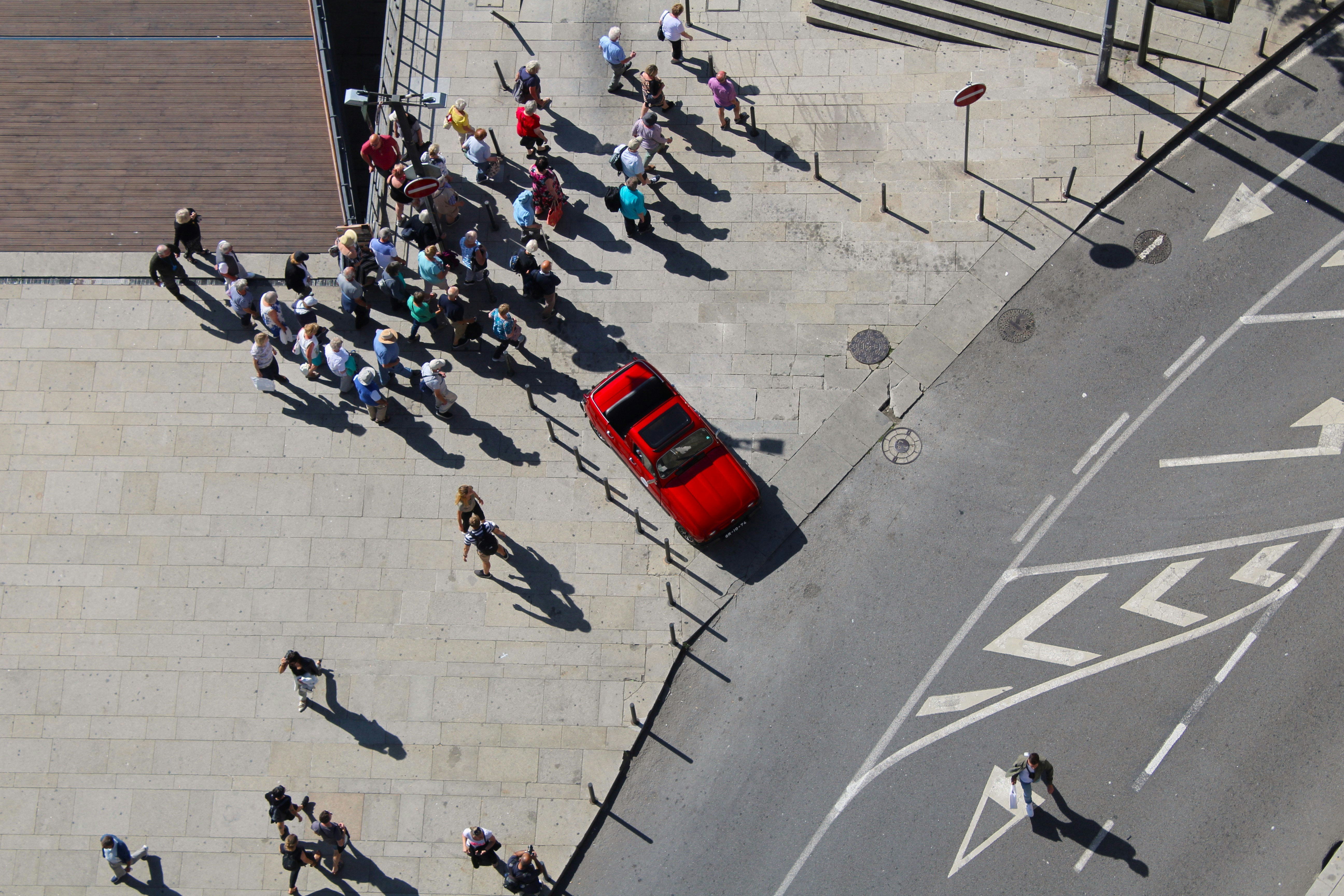 Top-down view of a red car and pedestrians at a city intersection casting long shadows.