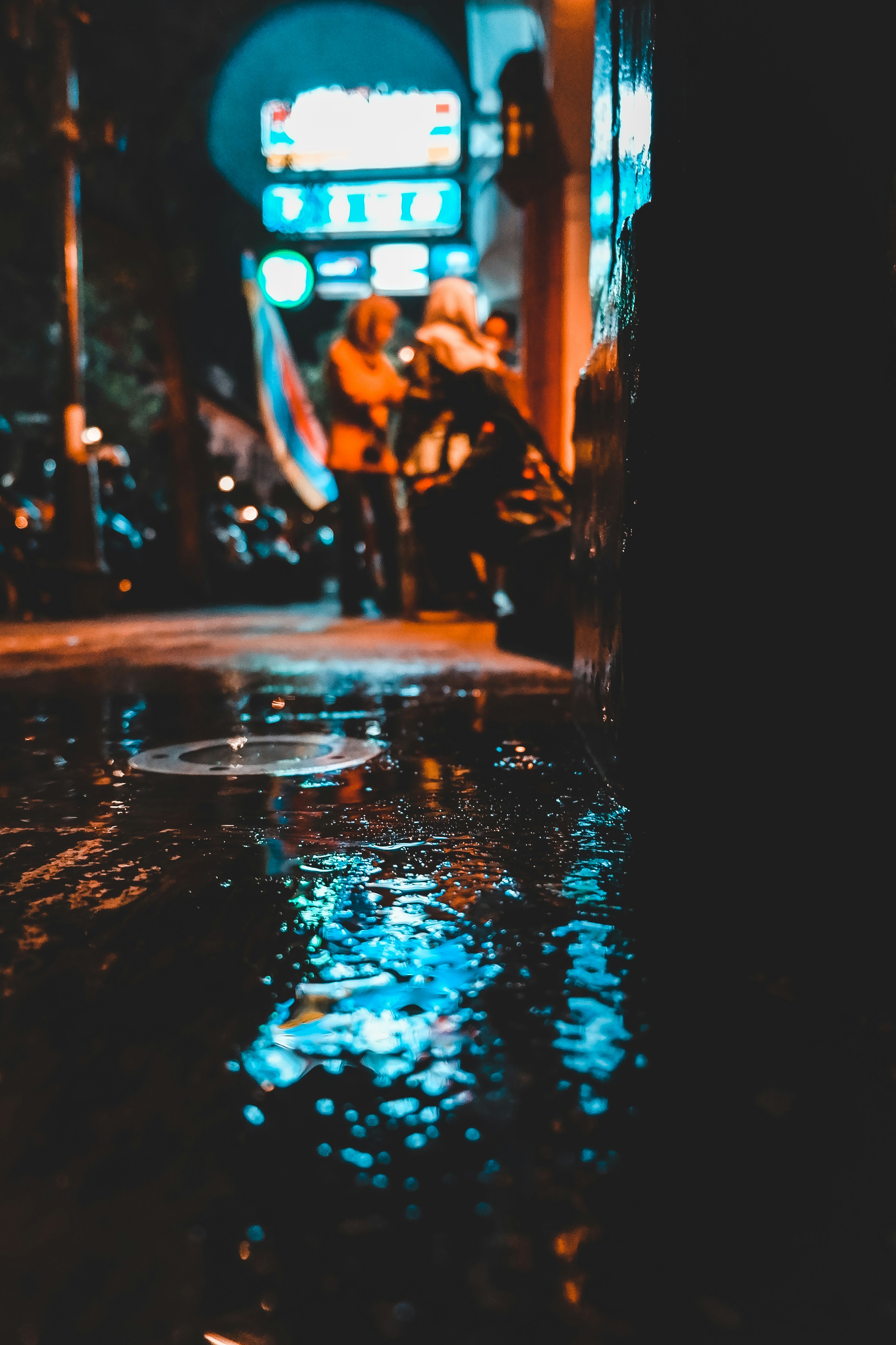 Night-time street photograph of a rain-soaked alley with neon signage reflecting on wet pavement. Blurred figures linger in the glow under storefront lights.