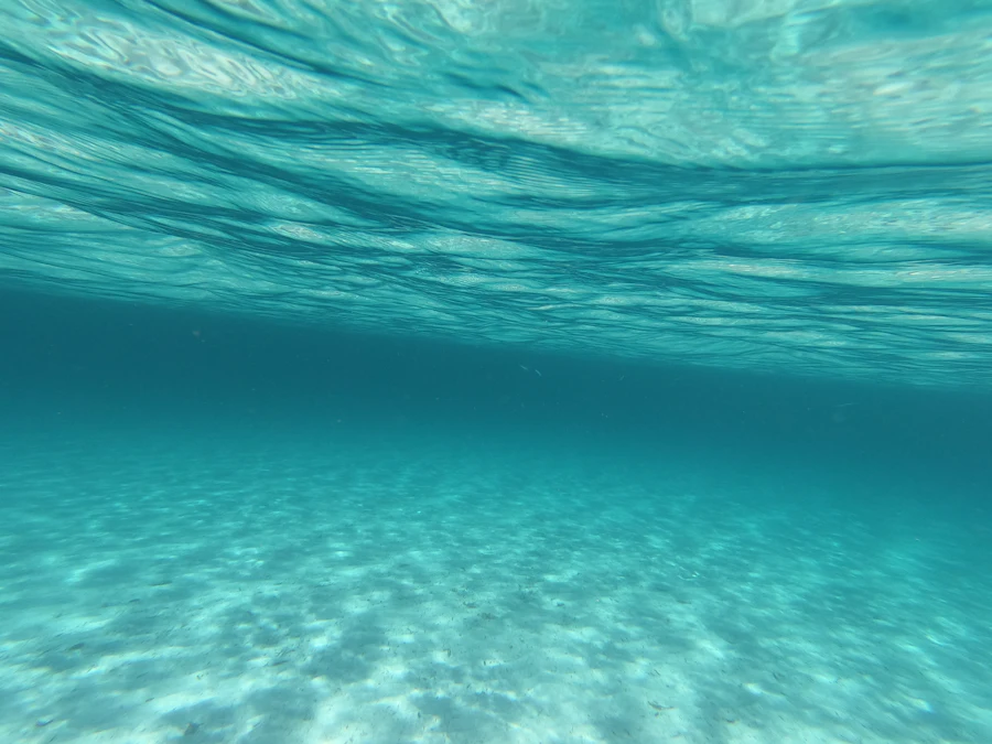 Underwater scene with rippled turquoise surface overhead and a sandy seafloor fading into blue distance. Sunlight creates soft moving patterns across the bottom.