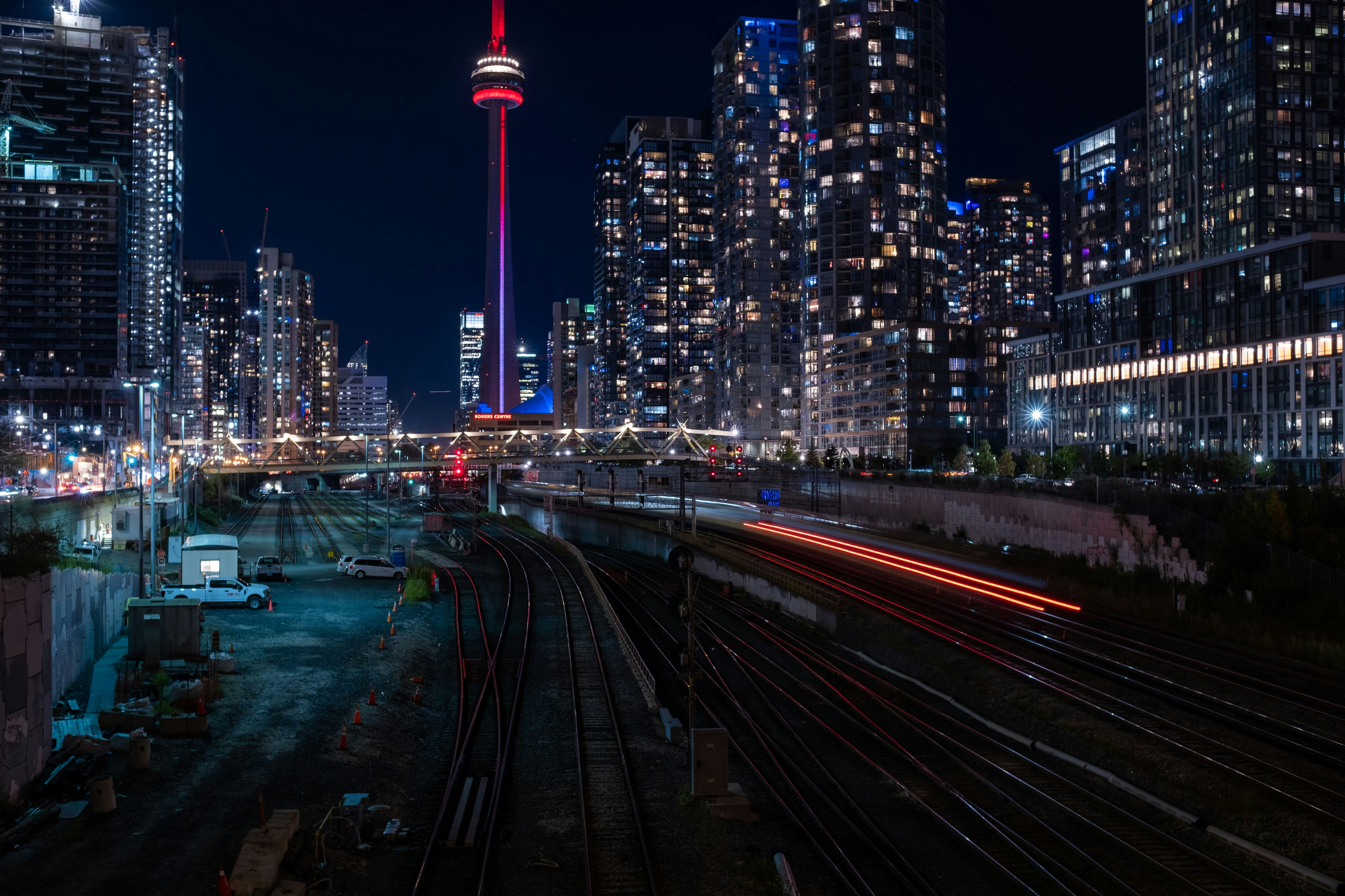The CN Tower illuminated in red amidst a backdrop of shimmering skyscrapers and train tracks at night.