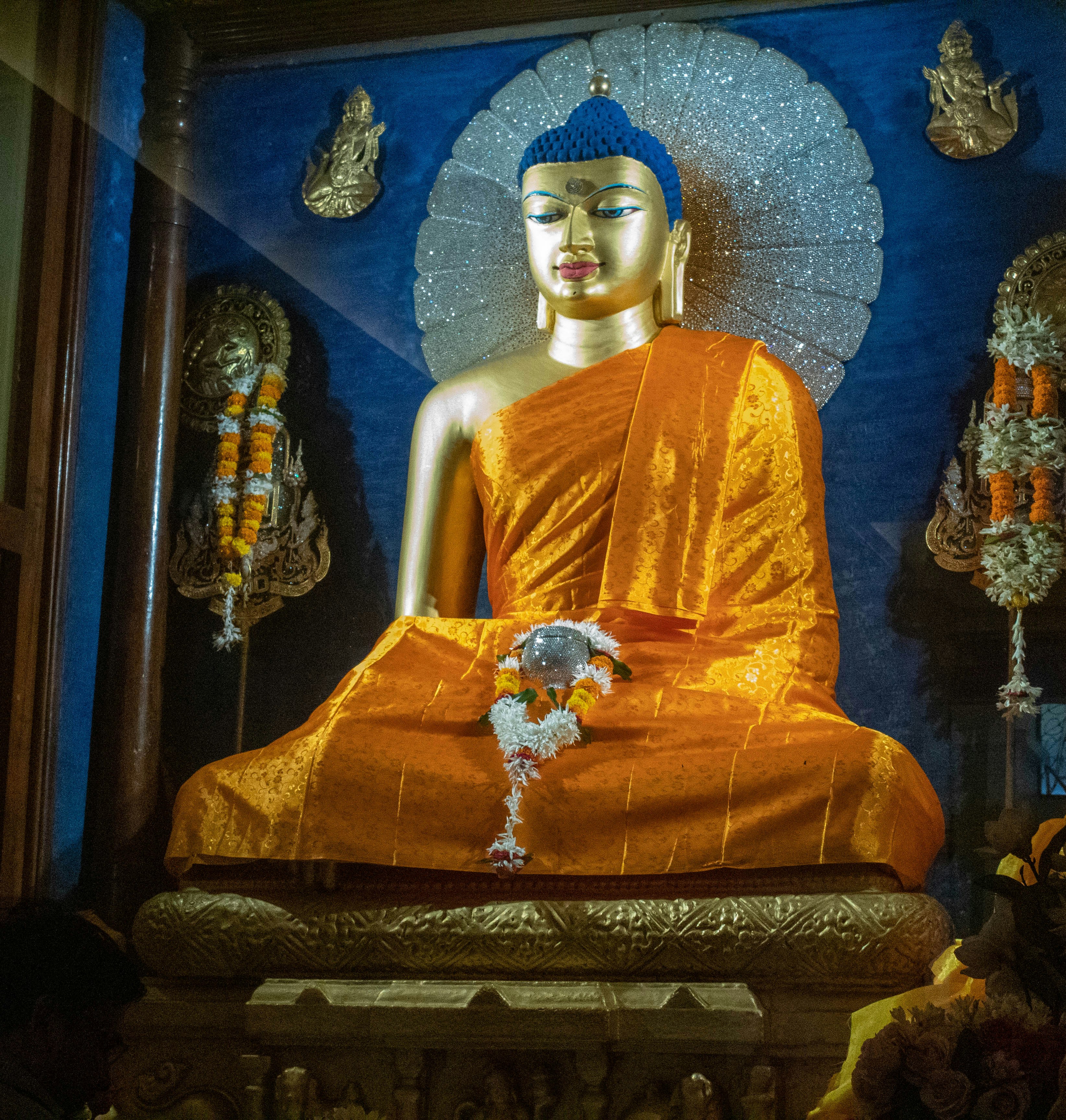 Golden statue of Buddha adorned with a vibrant orange robe, set against a blue backdrop with floral decorations and intricate carvings.