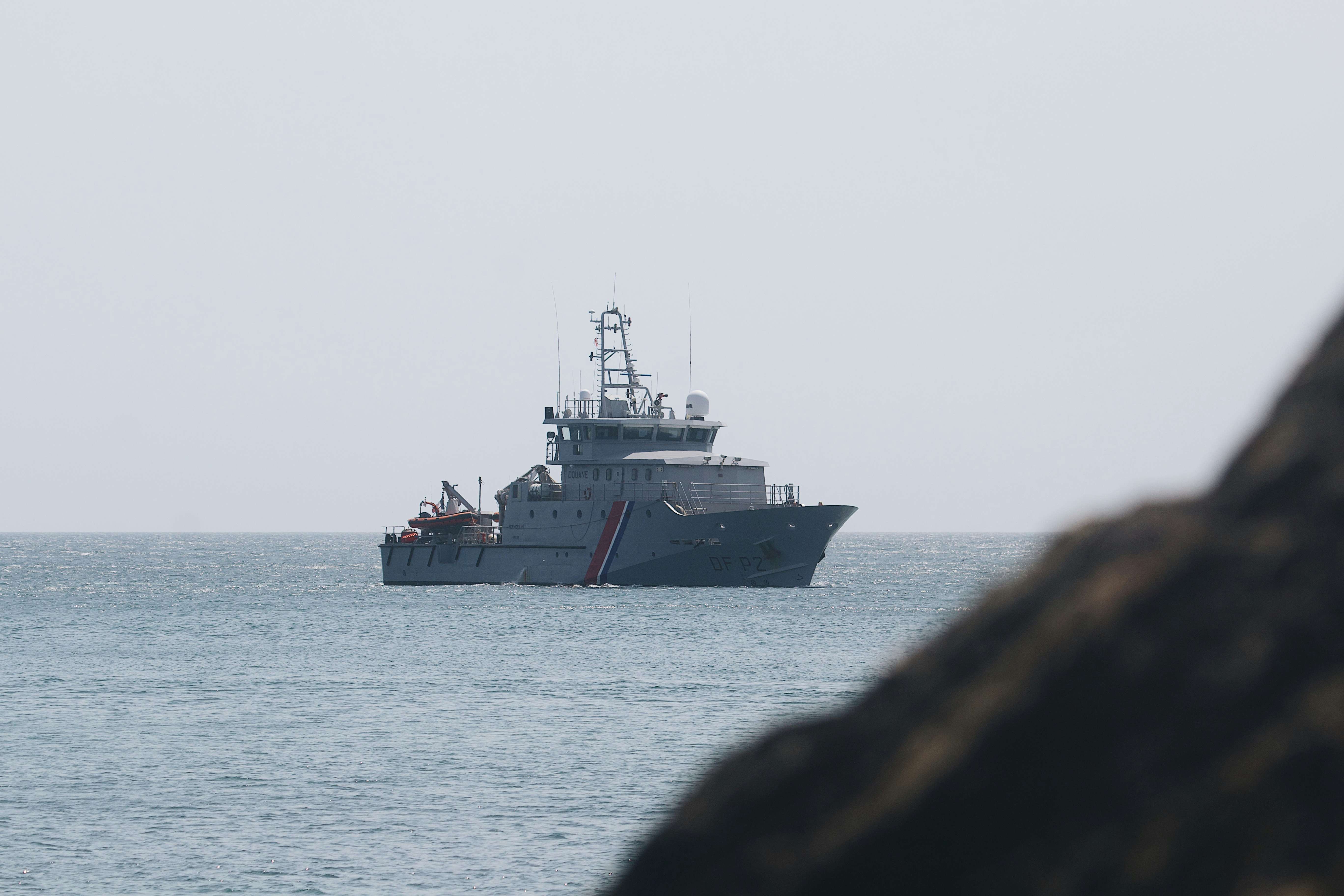 Navy ship cruising through calm seas with rocky foreground.