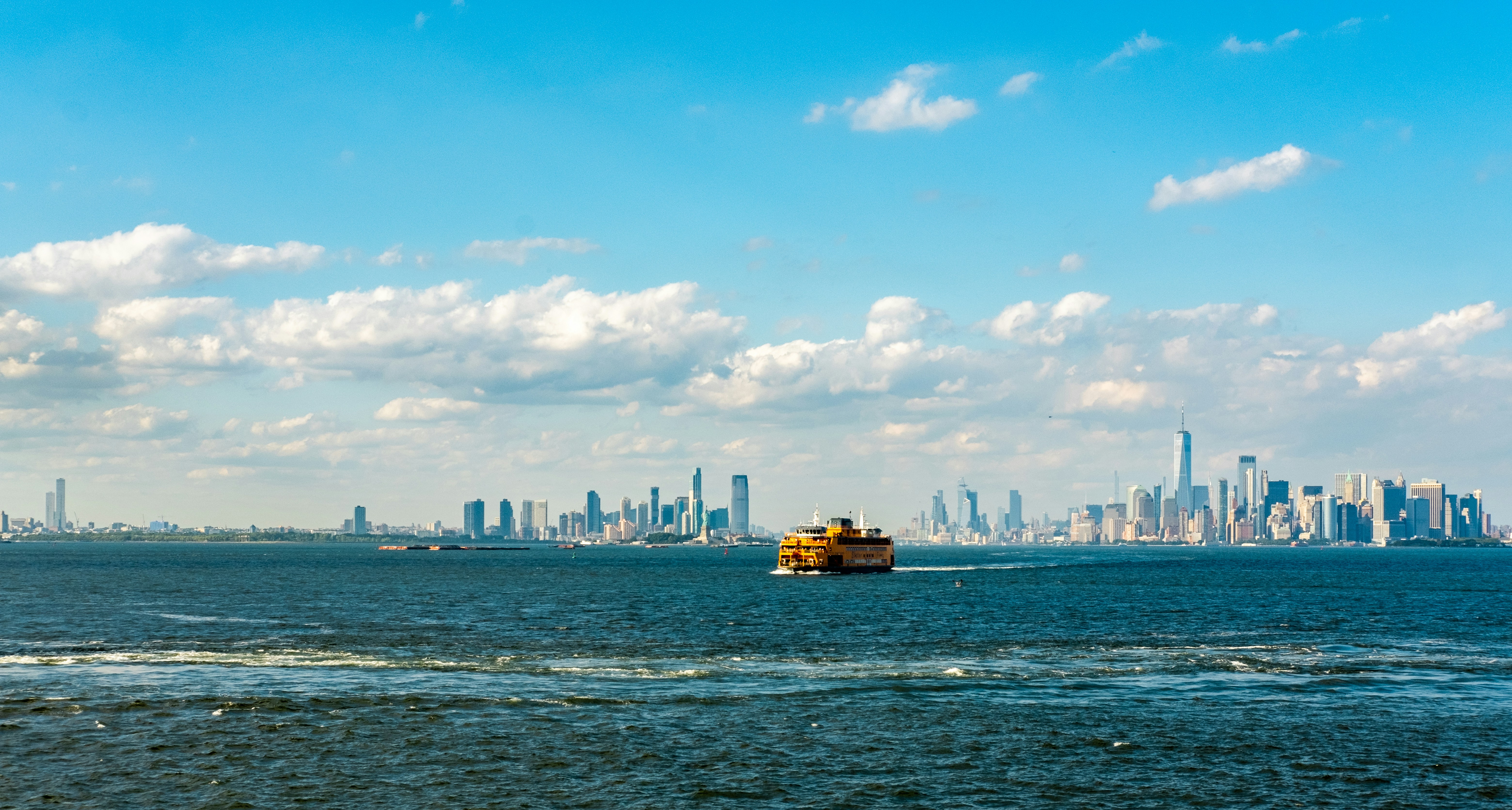 A ferry navigates the calm waters with a backdrop of a sprawling city skyline under a bright blue sky dotted with clouds.