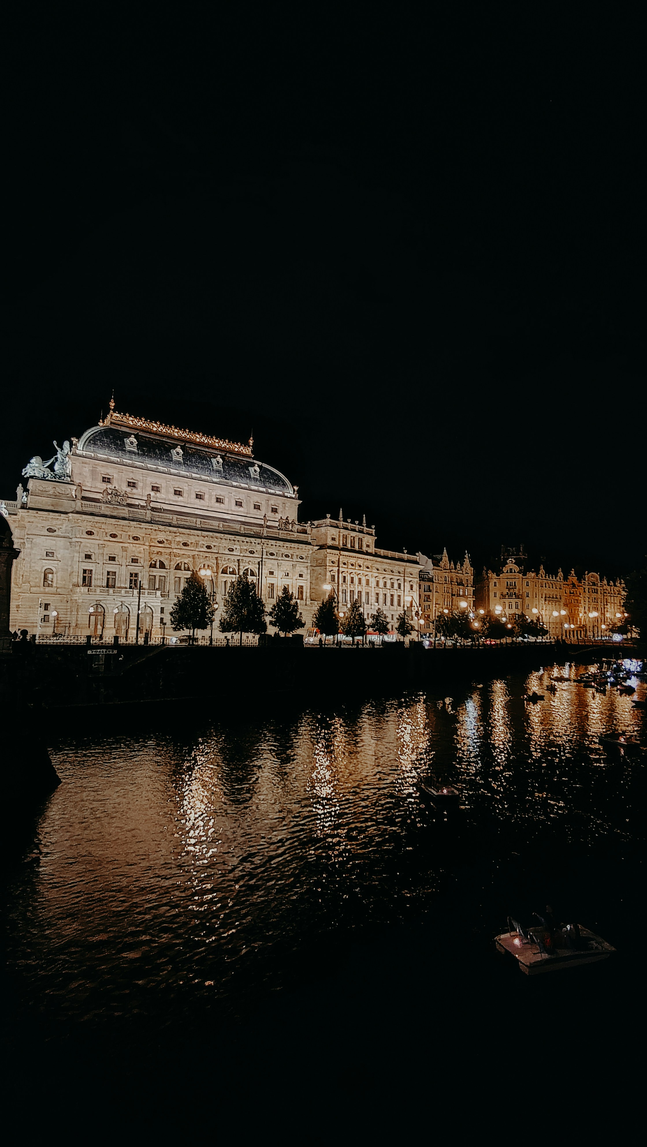 Historic riverside buildings glow with warm lights at night, their reflections shimmering on the dark water.