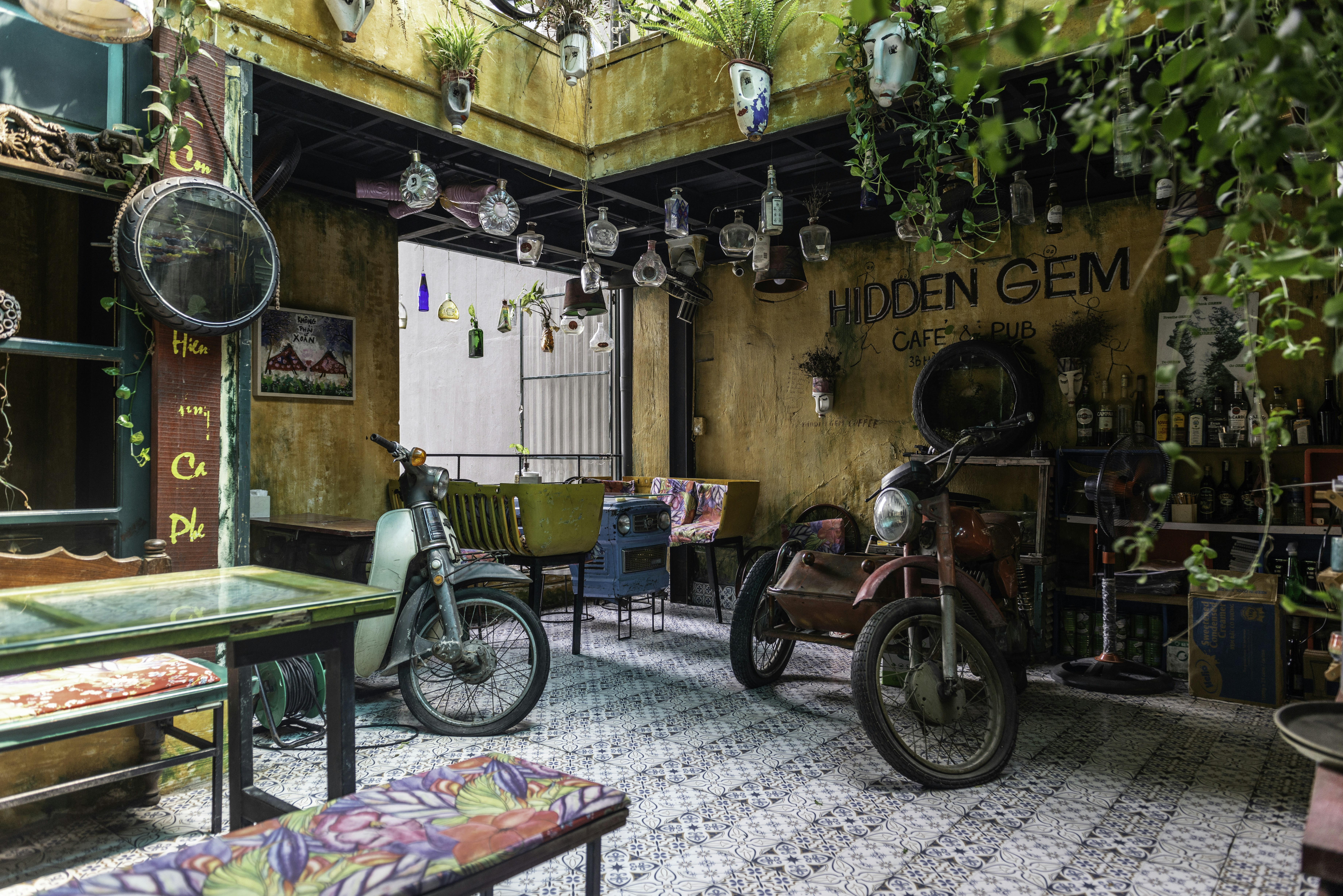 Two vintage motorcycles parked in a colorful, eclectic shop courtyard adorned with plants and retro decor.