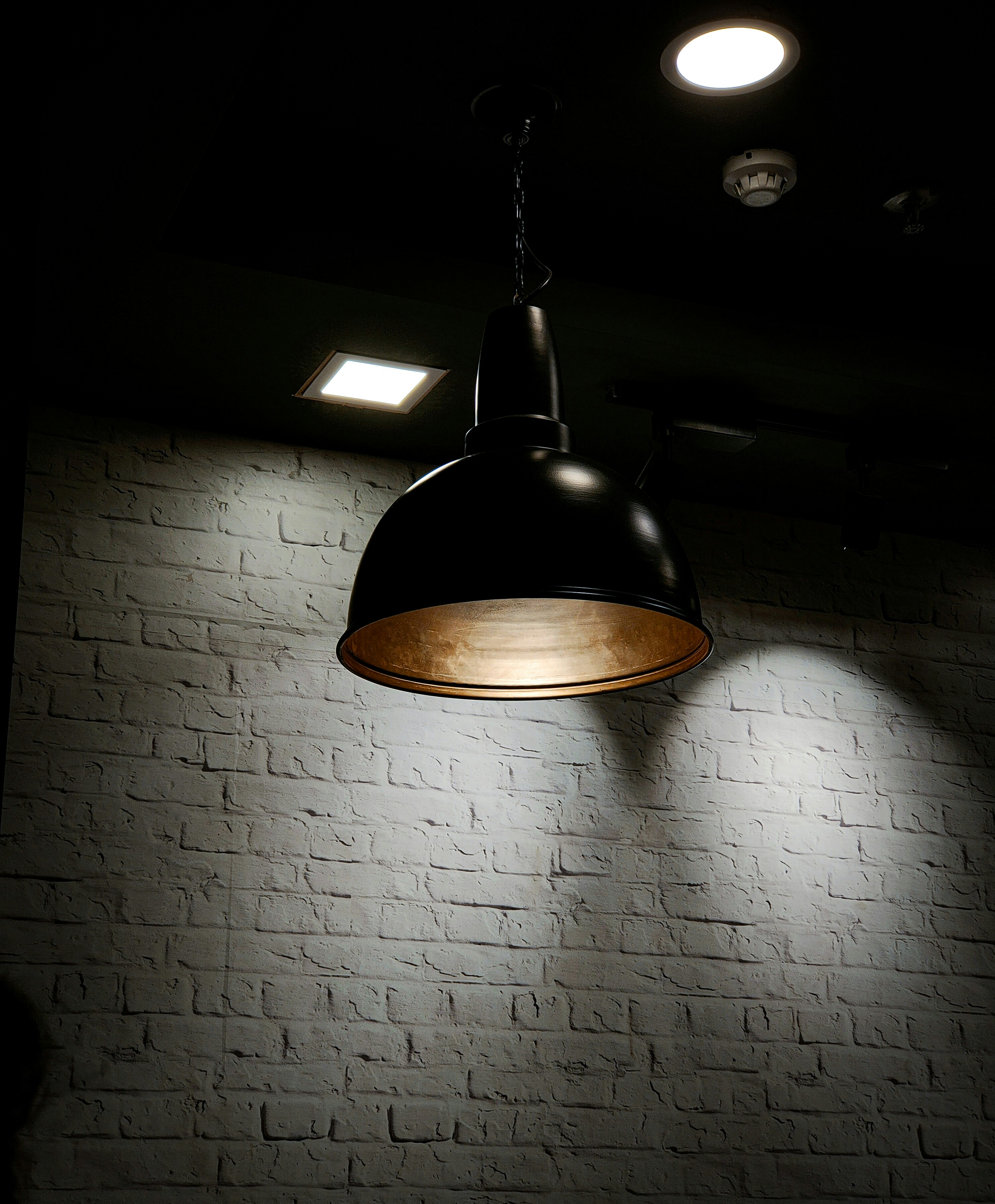Black industrial pendant hangs from a chain against a white brick wall. A warm copper interior emits a focused glow, creating a moody studio ambiance.