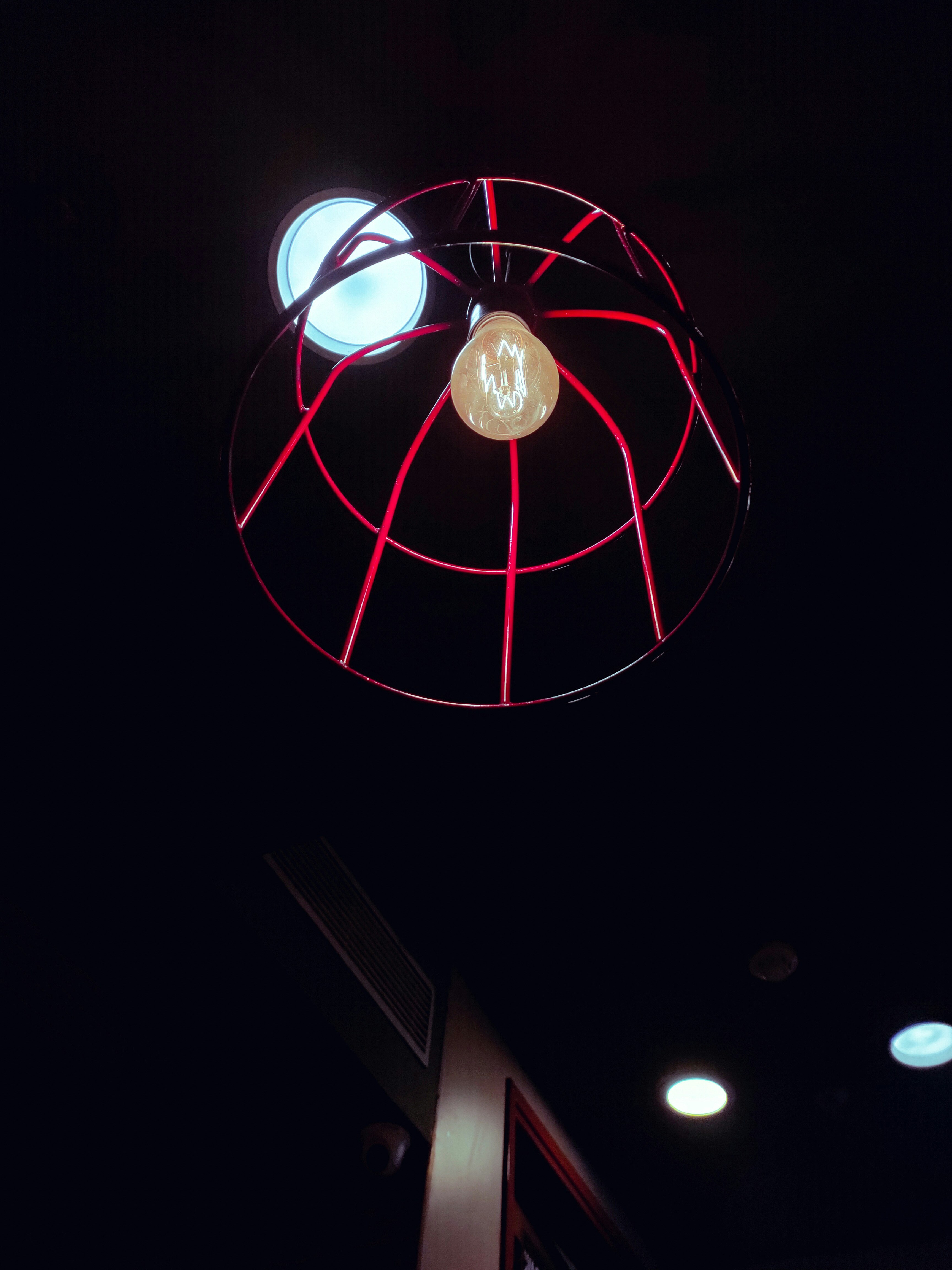 A modern pendant light fixture with a red wireframe design and a glowing bulb, suspended against a dark backdrop. The contrasting elements create a striking visual effect.