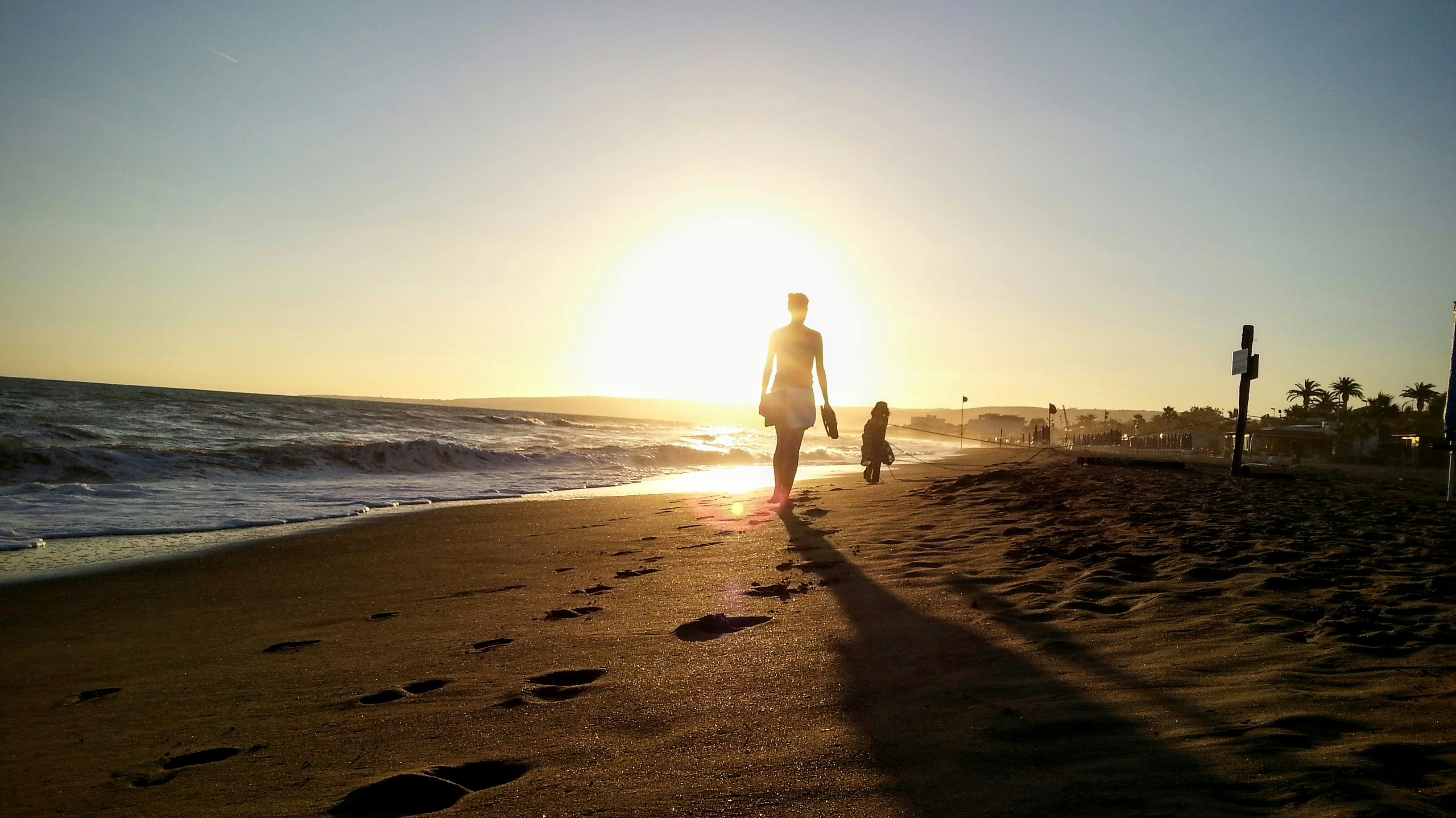 A person walks hand-in-hand with a child along a sunlit beach, with waves gently lapping at the shore. The sun sets in the background, creating a warm glow.