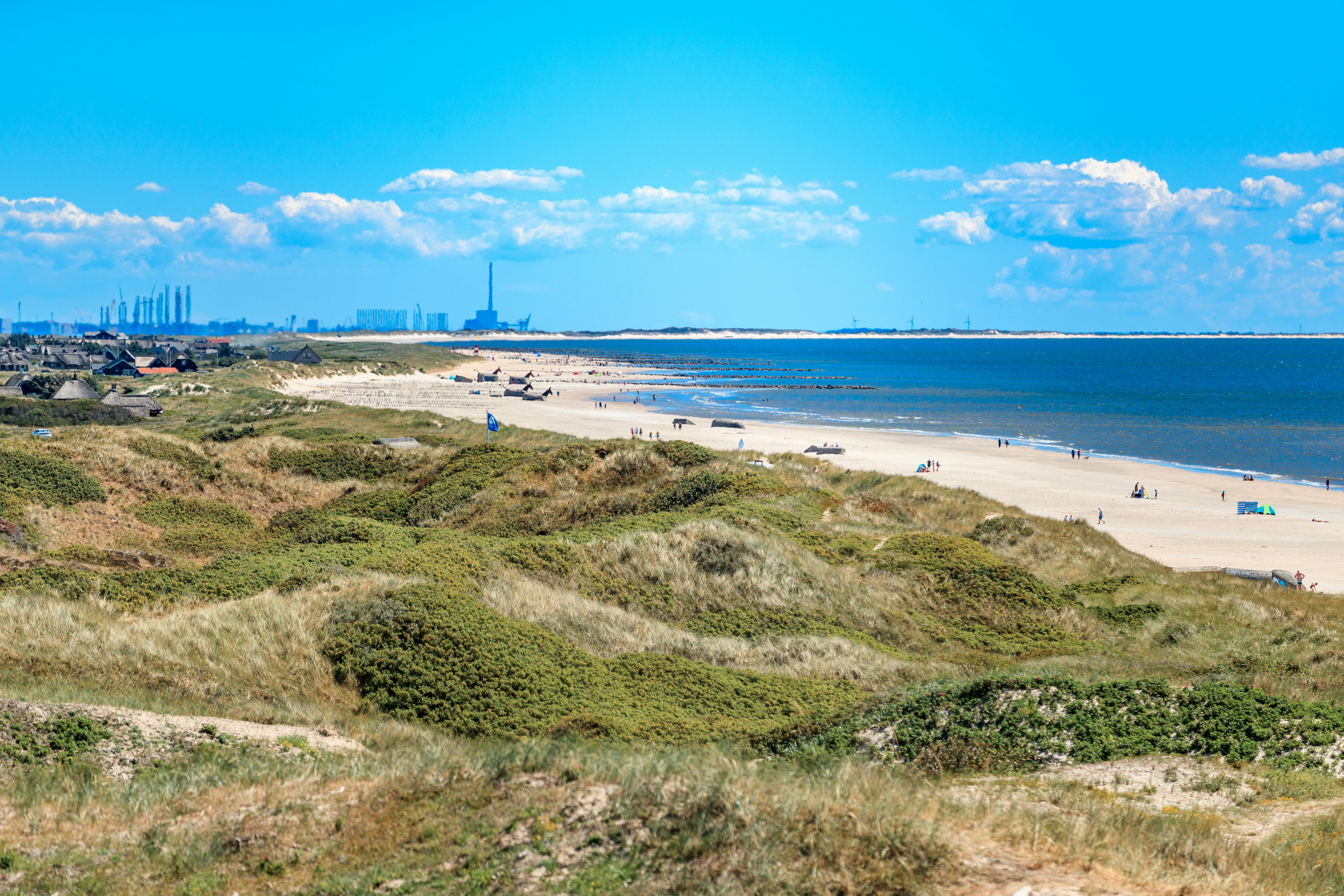 Rolling dunes meet a sandy beach under a bright blue sky, with distant industrial structures on the horizon.