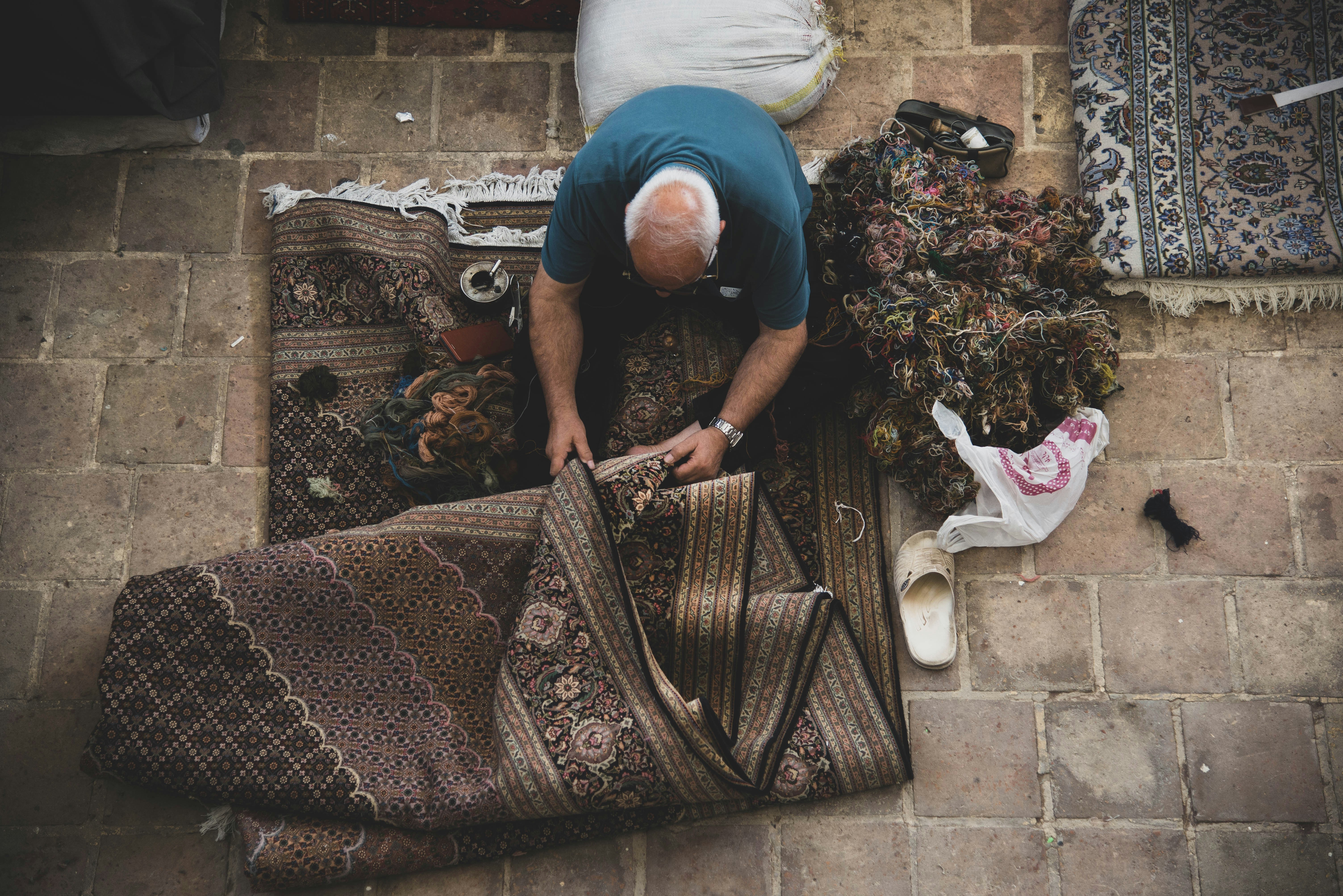 A craftsman meticulously repairs a patterned rug amidst vibrant yarns on a stone floor.