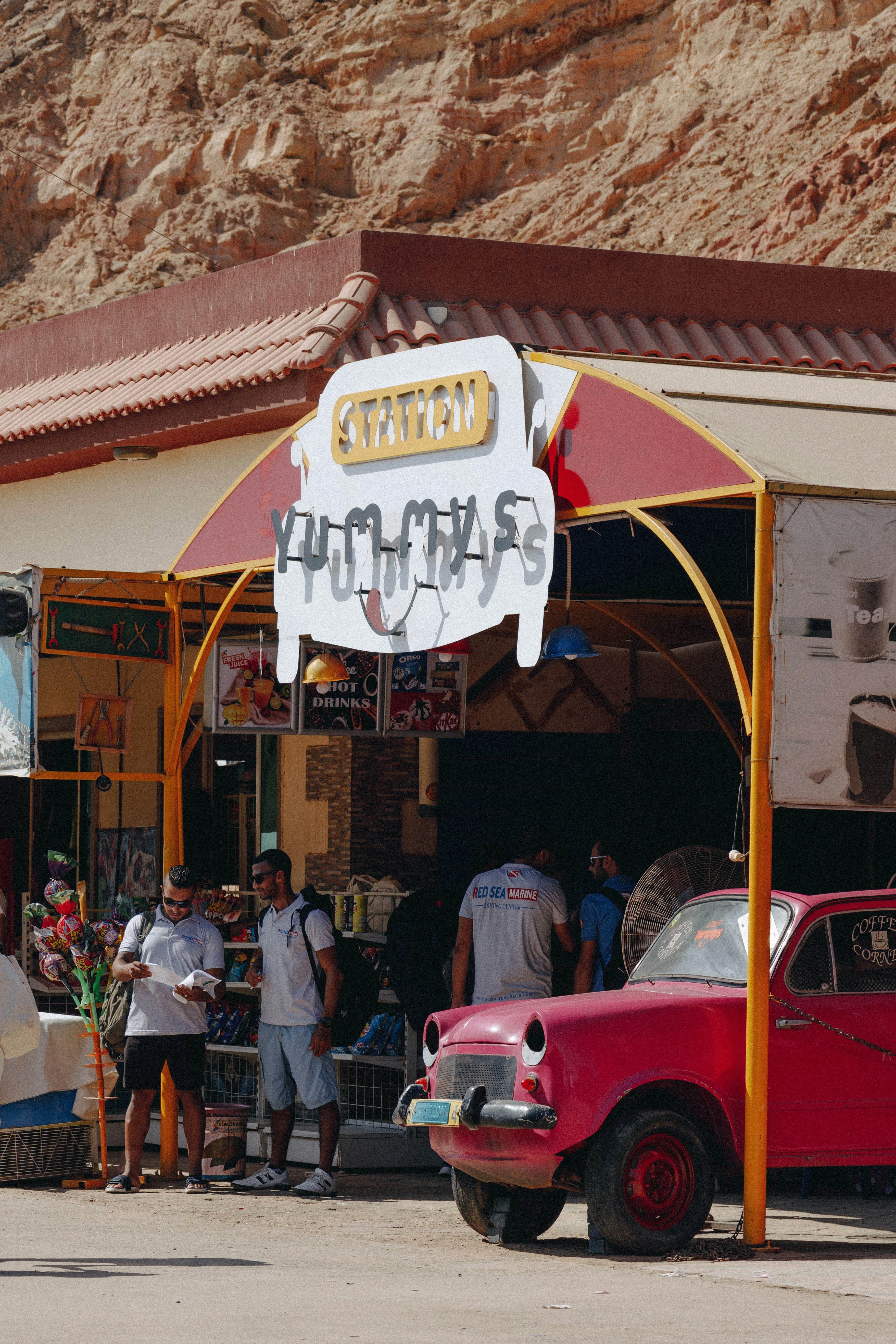 Colorful roadside station featuring a vintage red car and visitors engaging with local vendors. A lively atmosphere showcases the charm of community interaction.