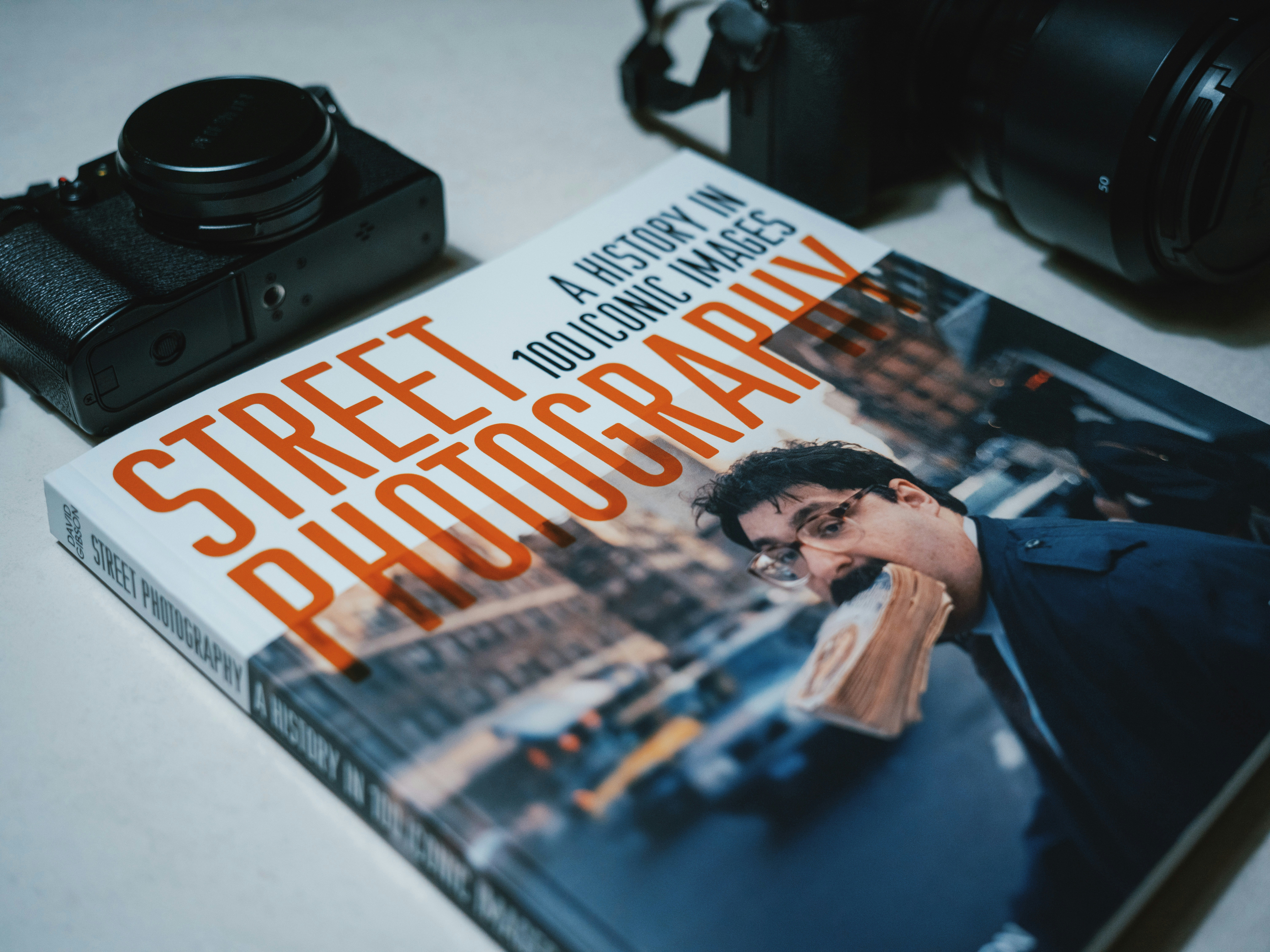 Open book titled 'Street Photography' featuring a man enjoying a sandwich, with cameras in the background. The scene captures the essence of street life and photography.